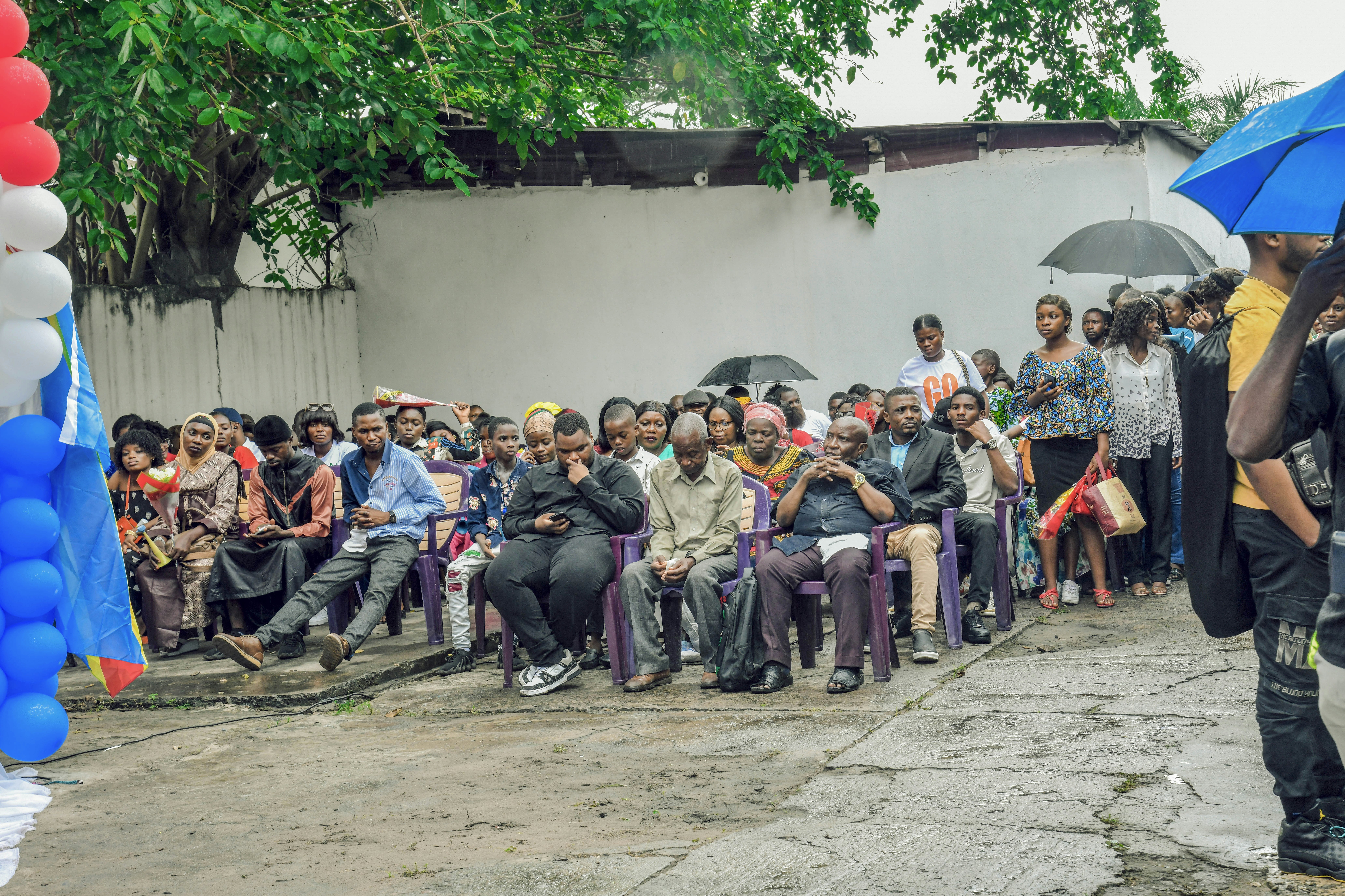 A diverse group of individuals seated in a semi-circle, some holding umbrellas, during a community event. The atmosphere reflects engagement and togetherness.