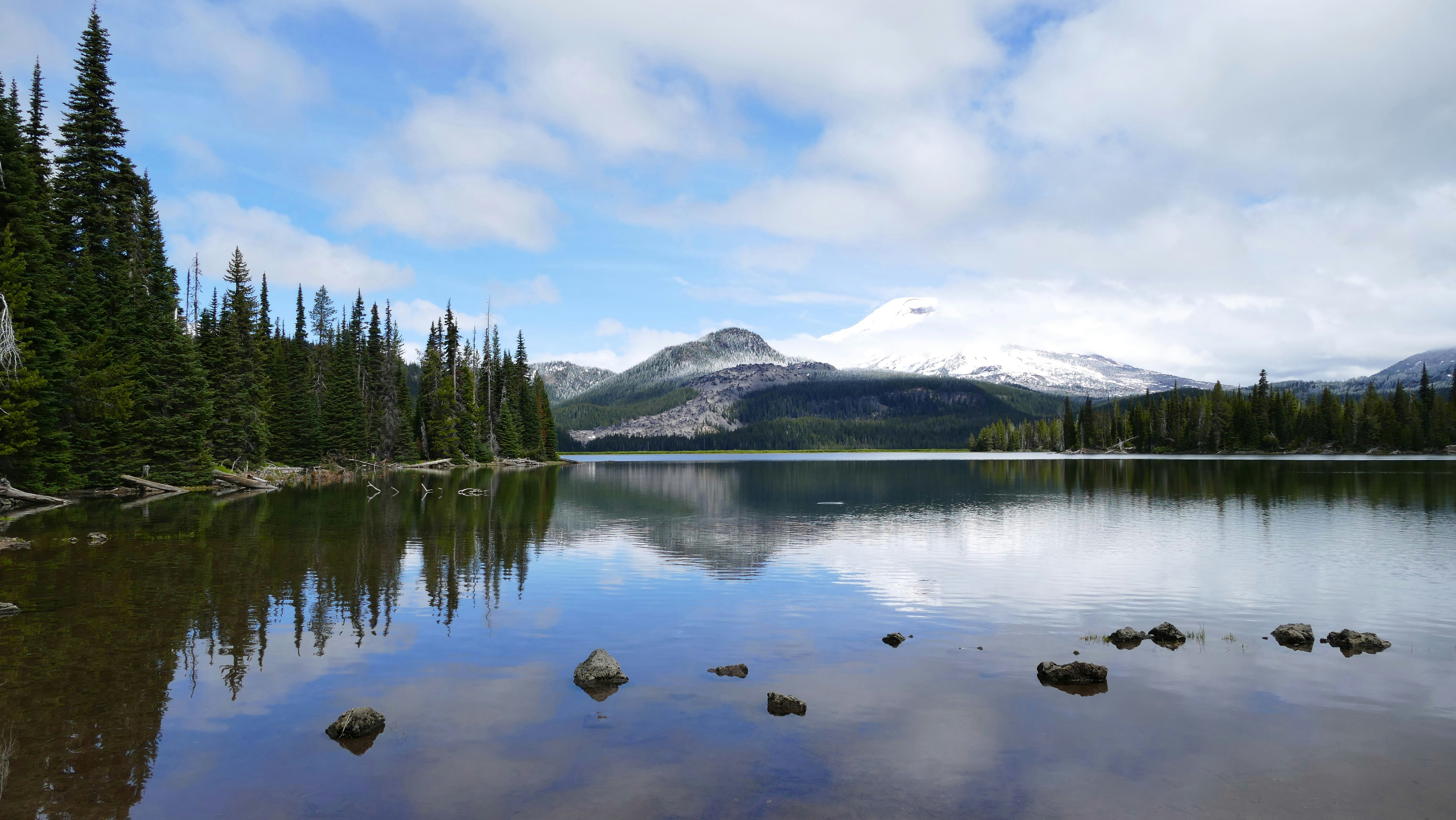 Calm lake reflecting surrounding trees and distant mountains beneath a cloudy sky.