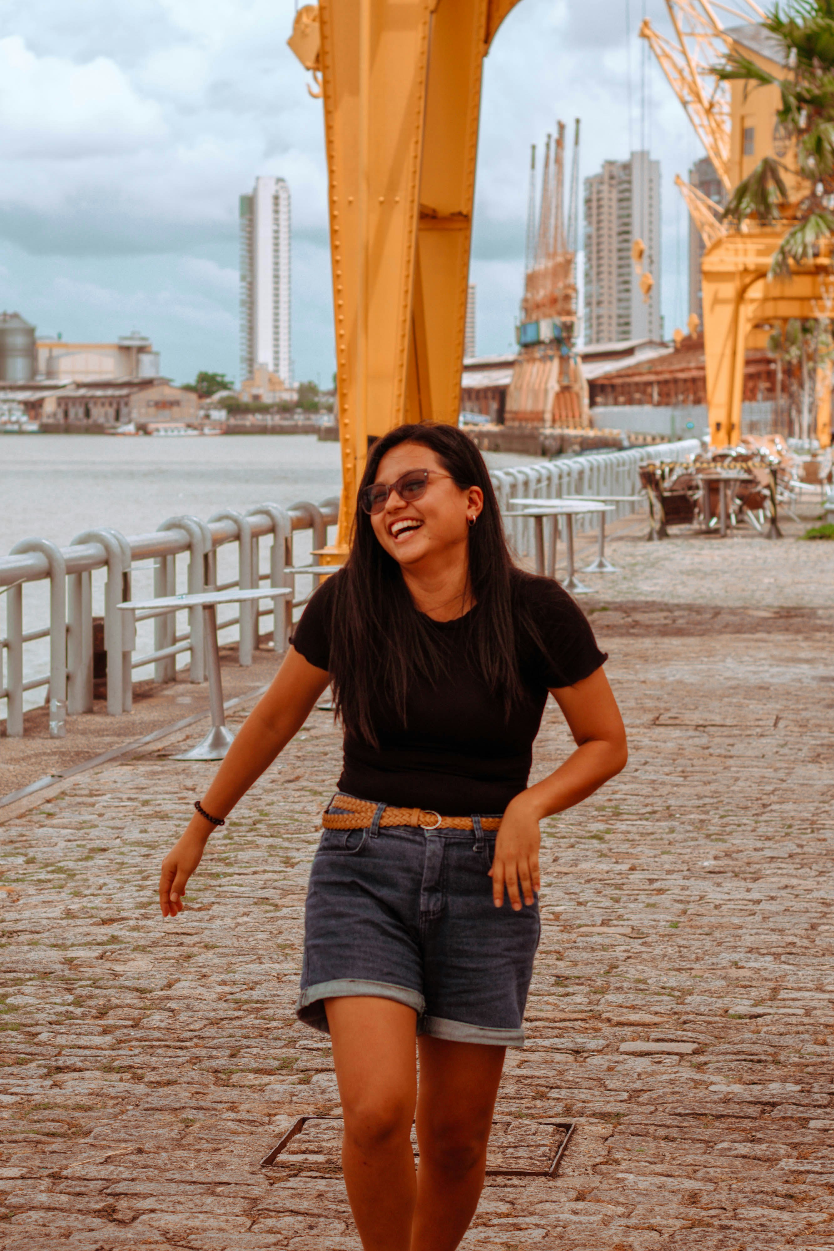 Woman laughing while walking on a cobblestone path beside a river with urban skyline in the background.