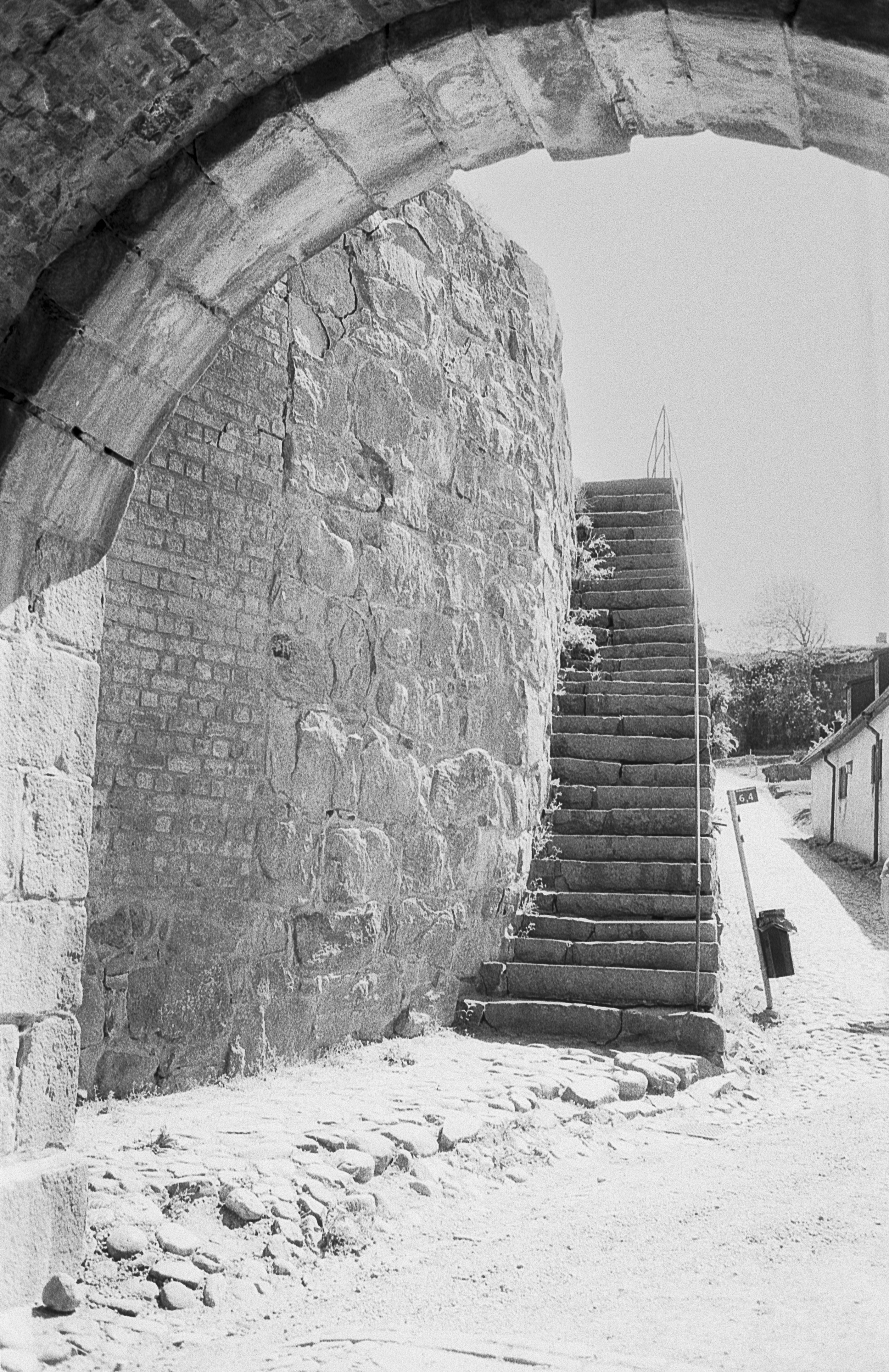 Black-and-white photograph of an arched stone structure with a broad staircase along a sunlit wall, leading toward a quiet street.