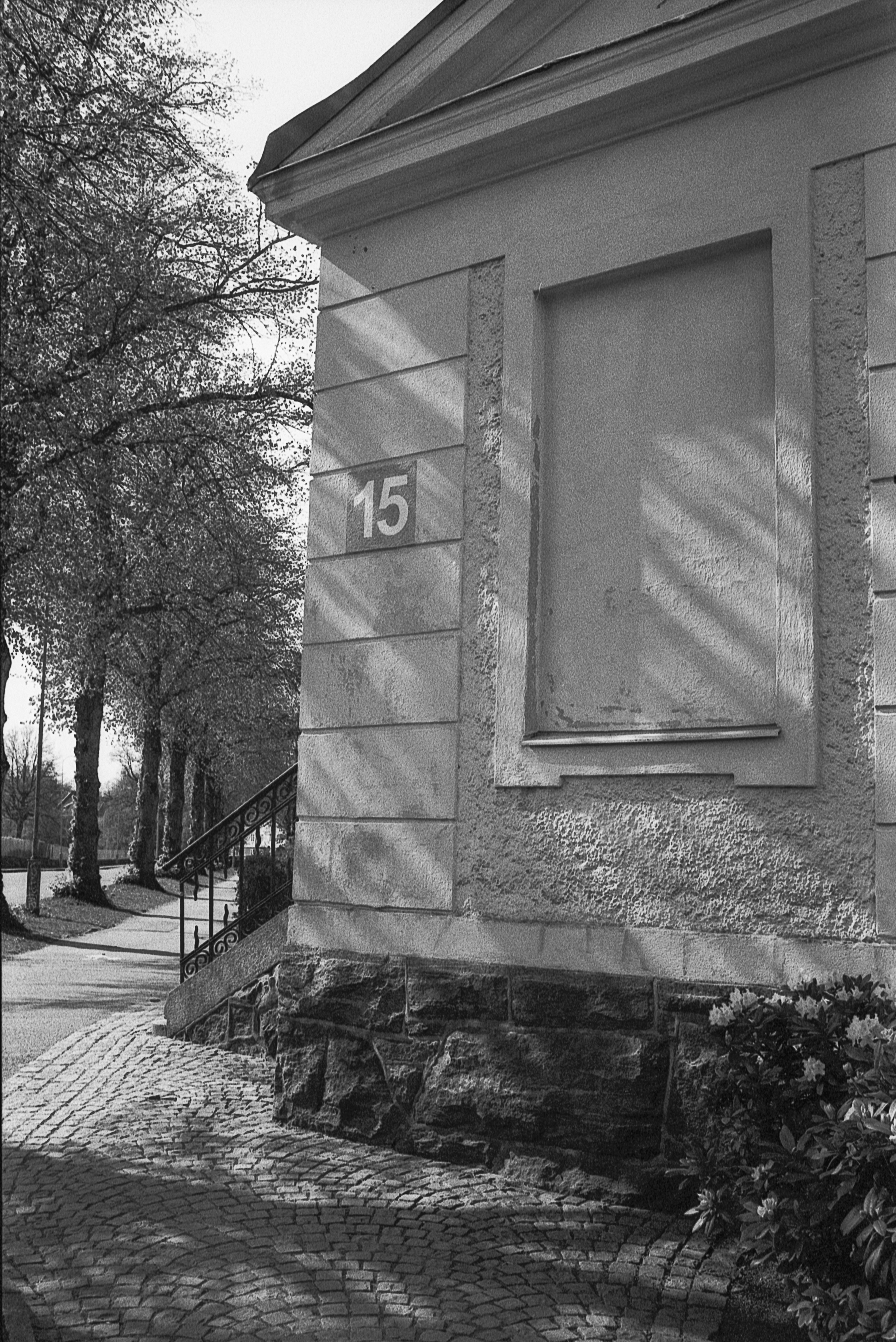 Monochrome street corner featuring a weathered wall with the number 15, a boarded window, and cobblestone paving; dappled tree shadows sweep across the facade.