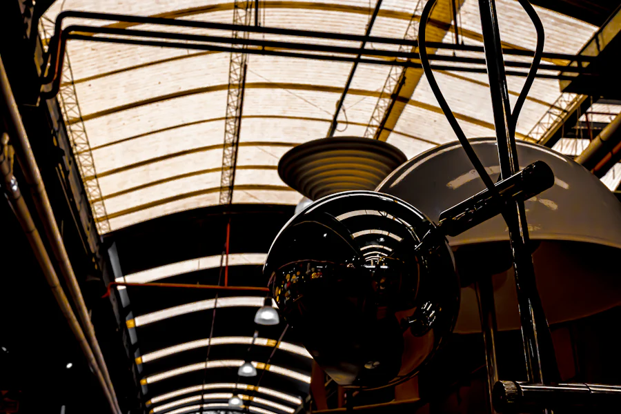 A glossy black mirrored sphere hangs beneath a vaulted industrial roof with repeating arches and warm skylights. Dark cables and structural beams frame the scene.