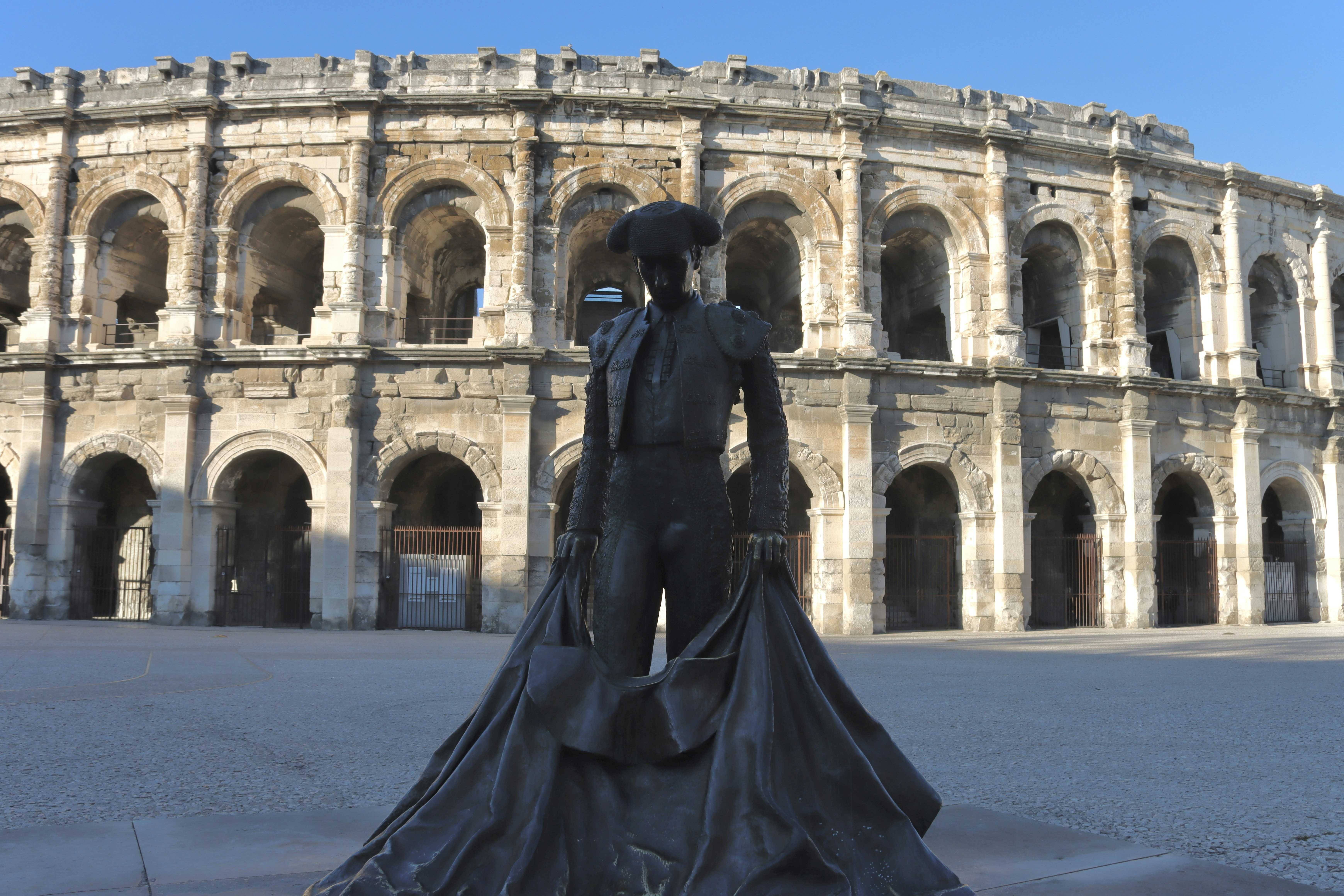 Bronze bullfighter statue stands before the historic Arena of Nîmes under a clear blue sky.