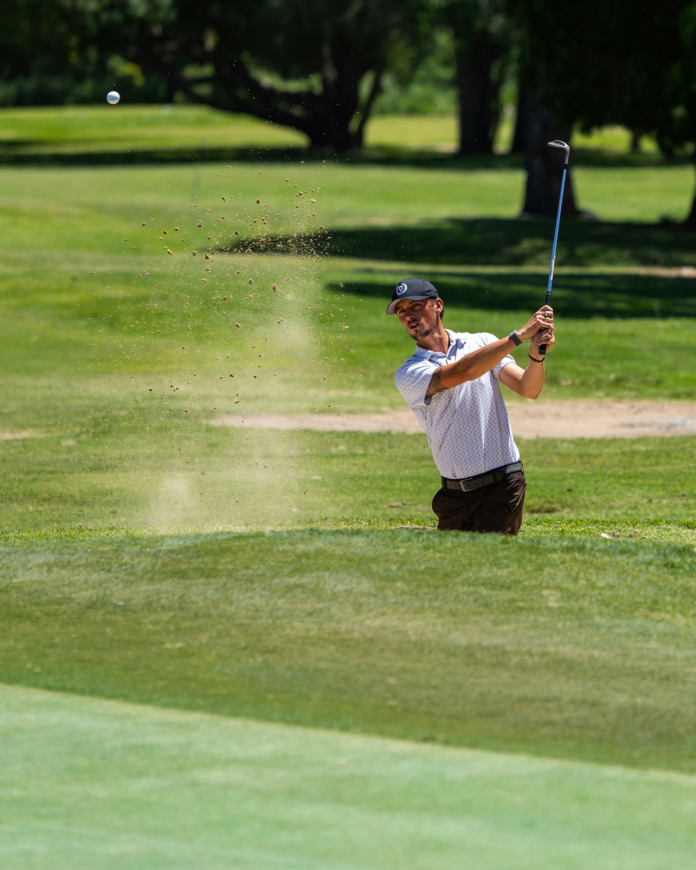 Golfer in a white polo executes a full swing on a sunlit golf course. Sand and turf erupt as the ball sails through the frame, highlighting motion and precision.