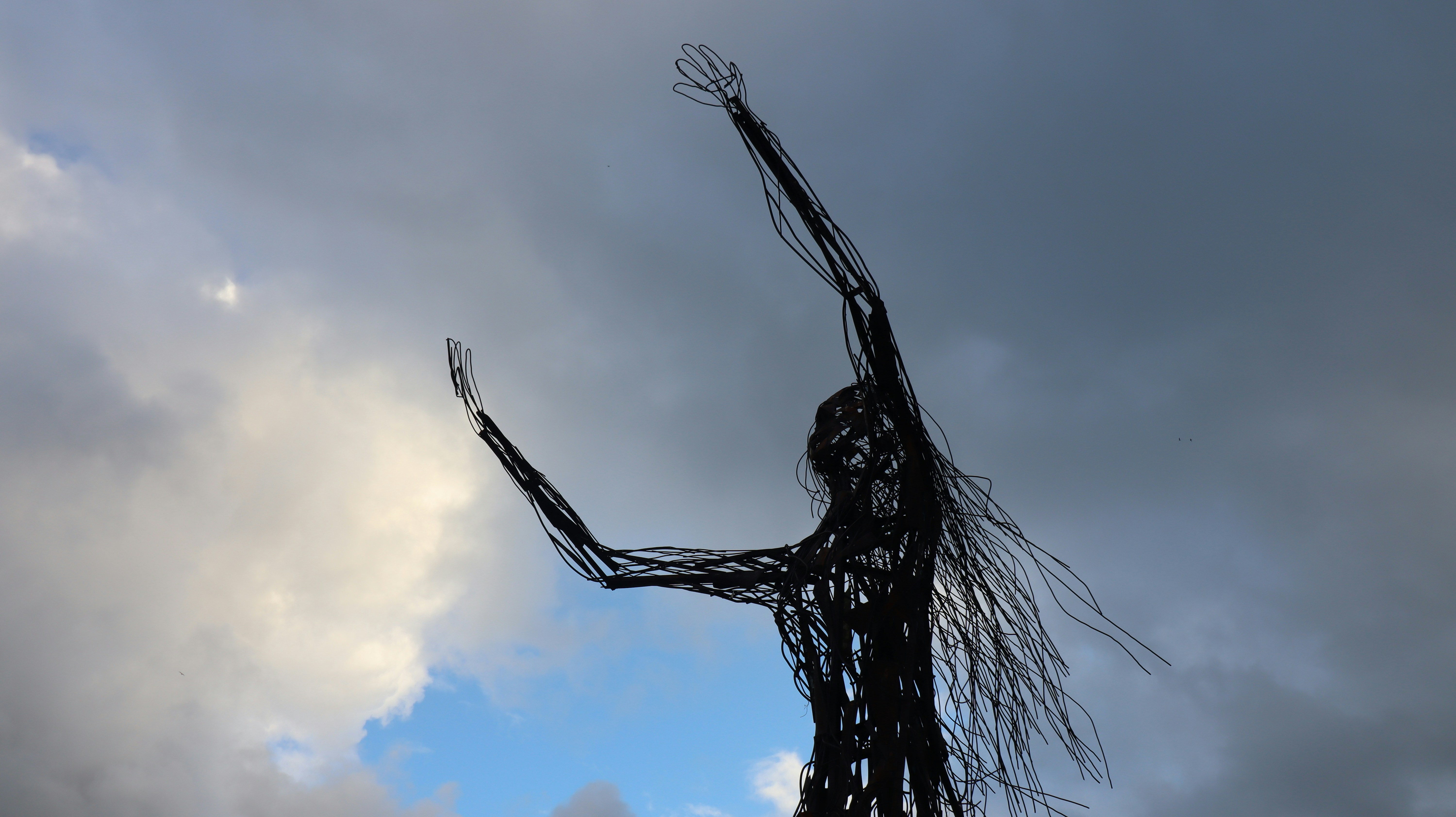 Metal sculpture of a figure reaching upward against a dramatic cloudy sky.