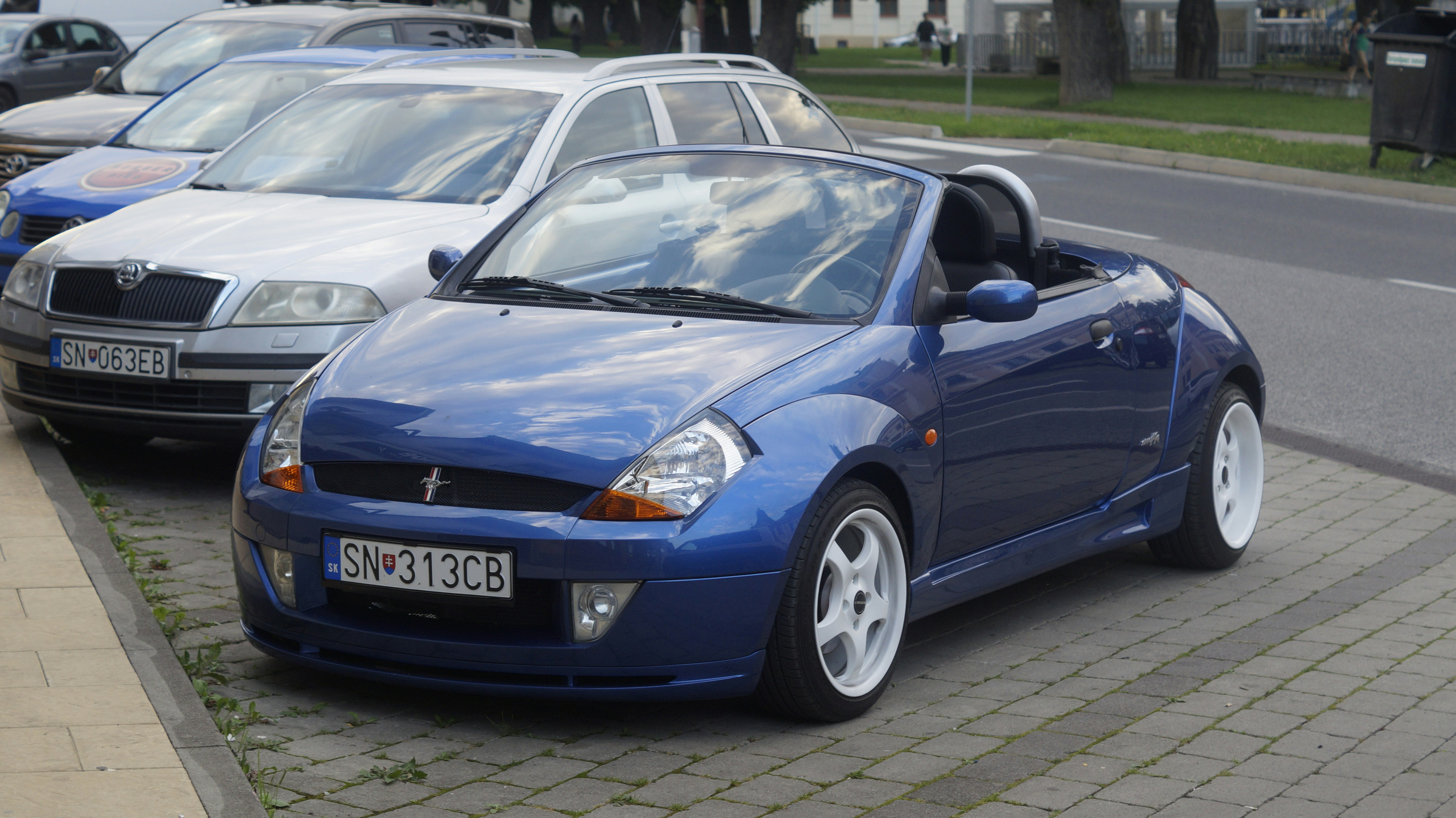 Blue convertible parked on a city street, surrounded by other vehicles on a sunny day.