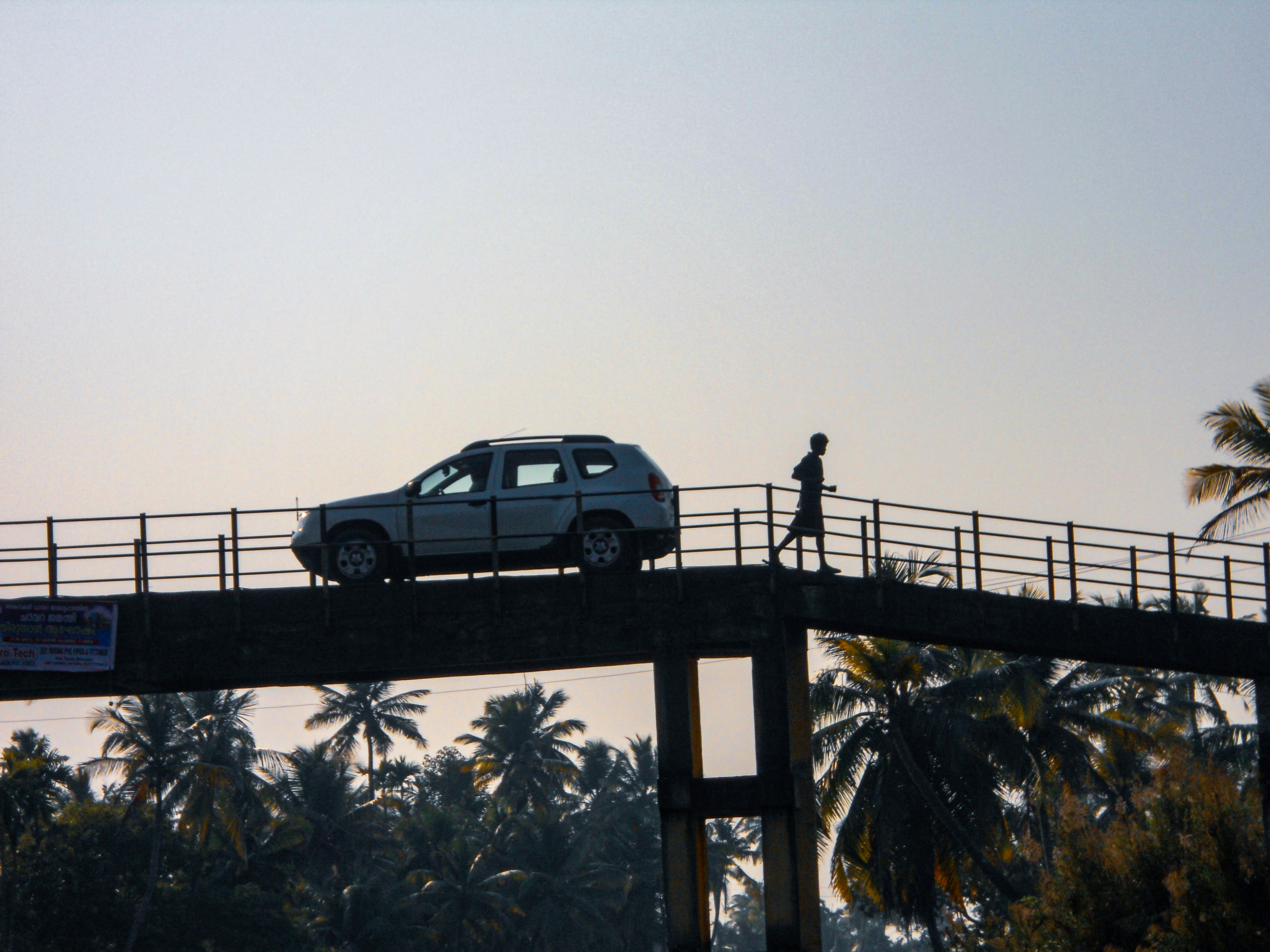 Silhouetted SUV on a narrow bridge with a lone walker against a palm-lined backdrop at dusk.