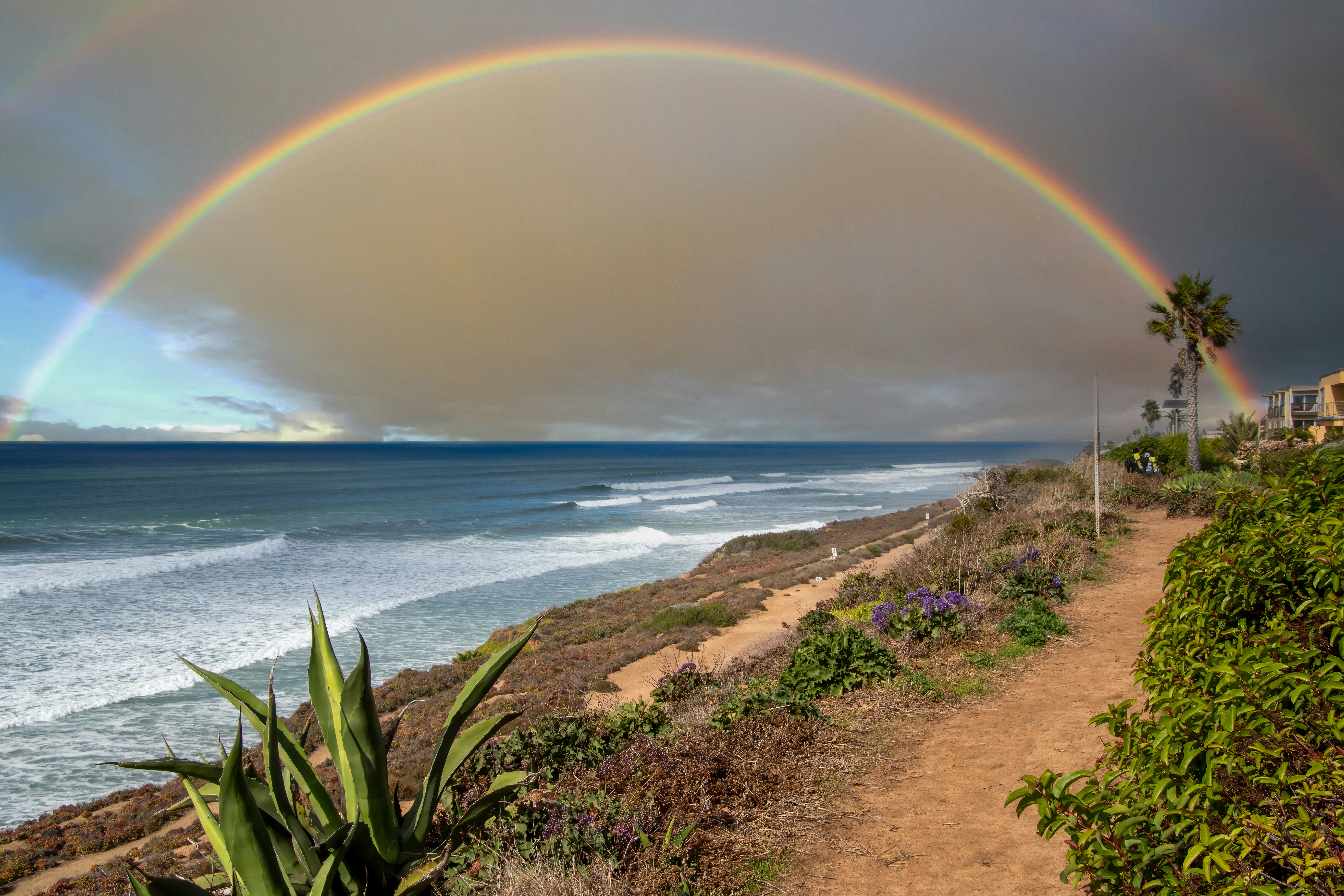 Two rainbows arc over a rugged cliffside path leading to a tranquil ocean under a dramatic sky.