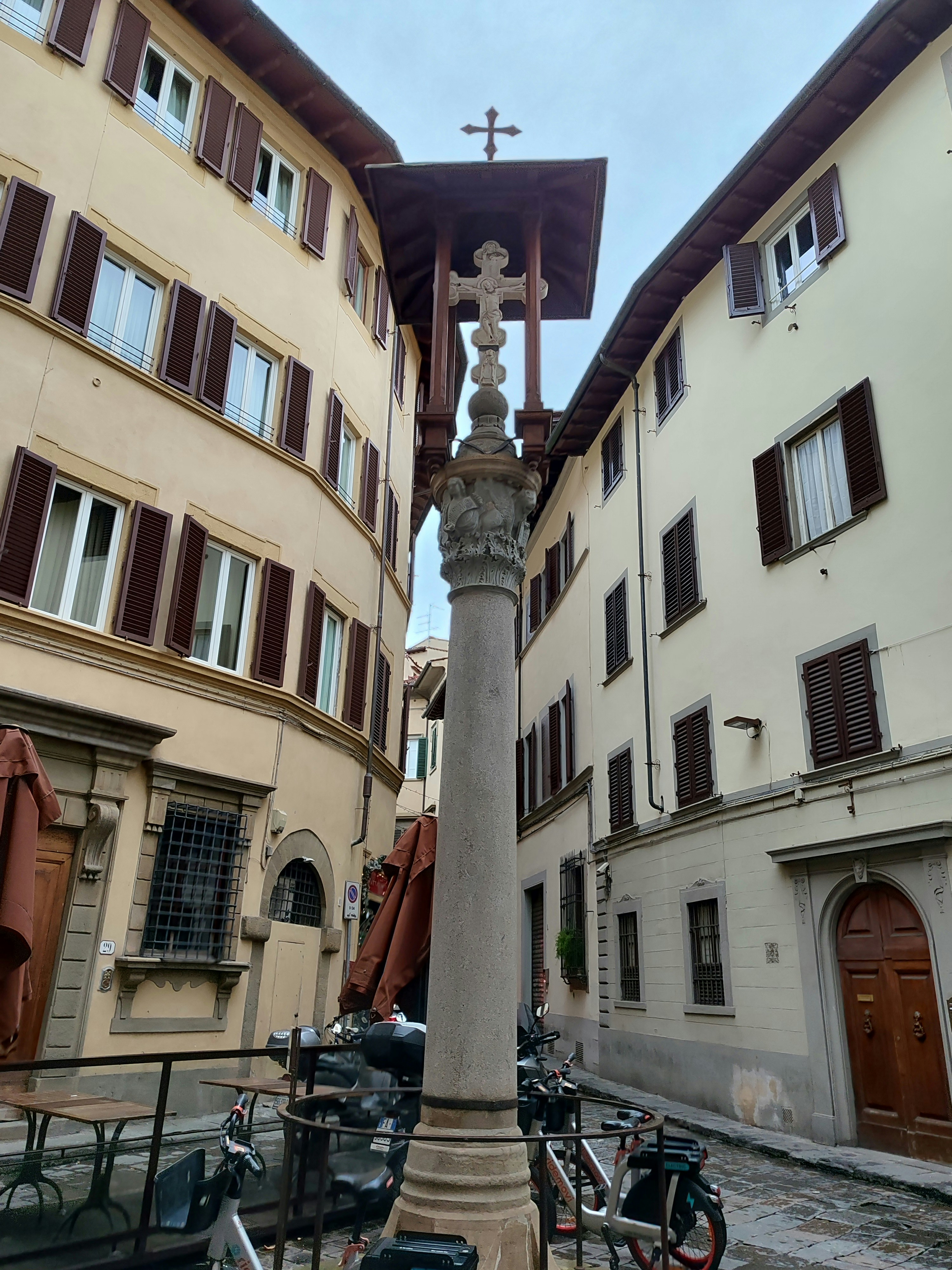 Stone cross atop a decorative pillar in a quaint European street between traditional buildings.