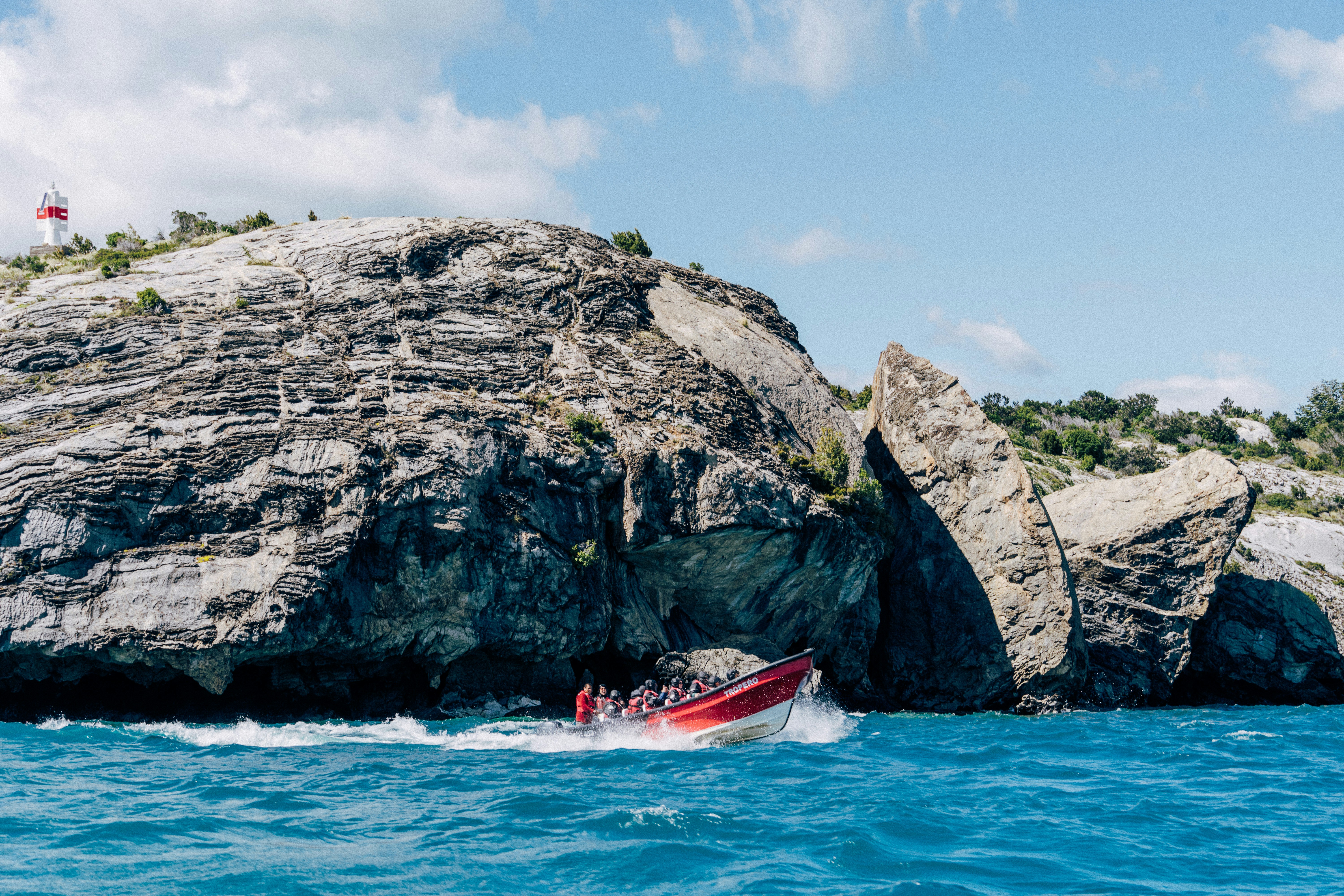 A vibrant red boat cuts through the striking turquoise water, set against the rugged backdrop of towering, textured cliffs. The scene is illuminated by bright daylight, casting dynamic shadows that enhance the dramatic rock formations. The image captures a sense of adventure and natural beauty, accentuated by the vivid contrast between the rich colors of the sea and the earthy tones of the rocks.