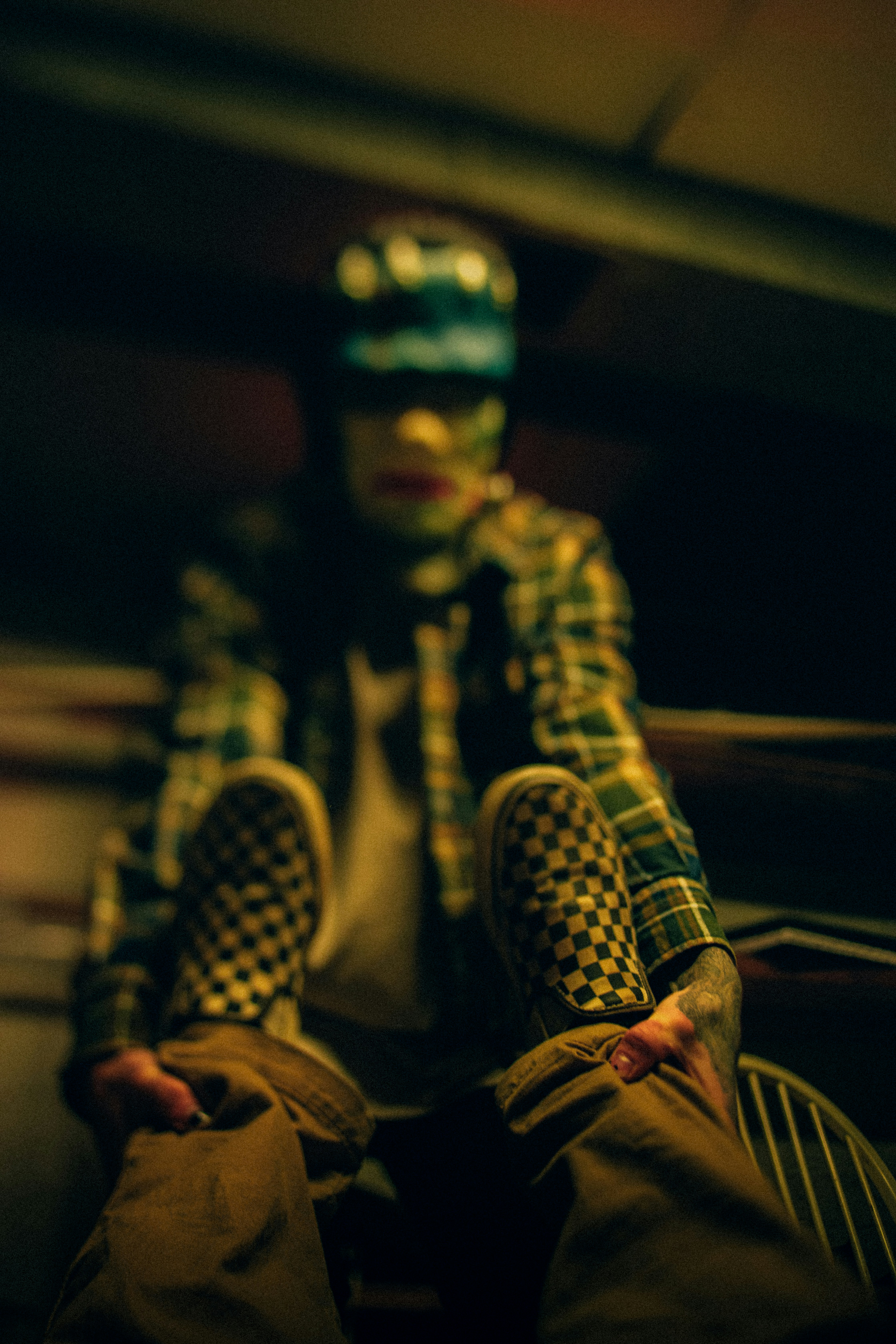A figure in a playful mask and checkered shoes reaches out, creating an intriguing perspective in a dimly lit space.