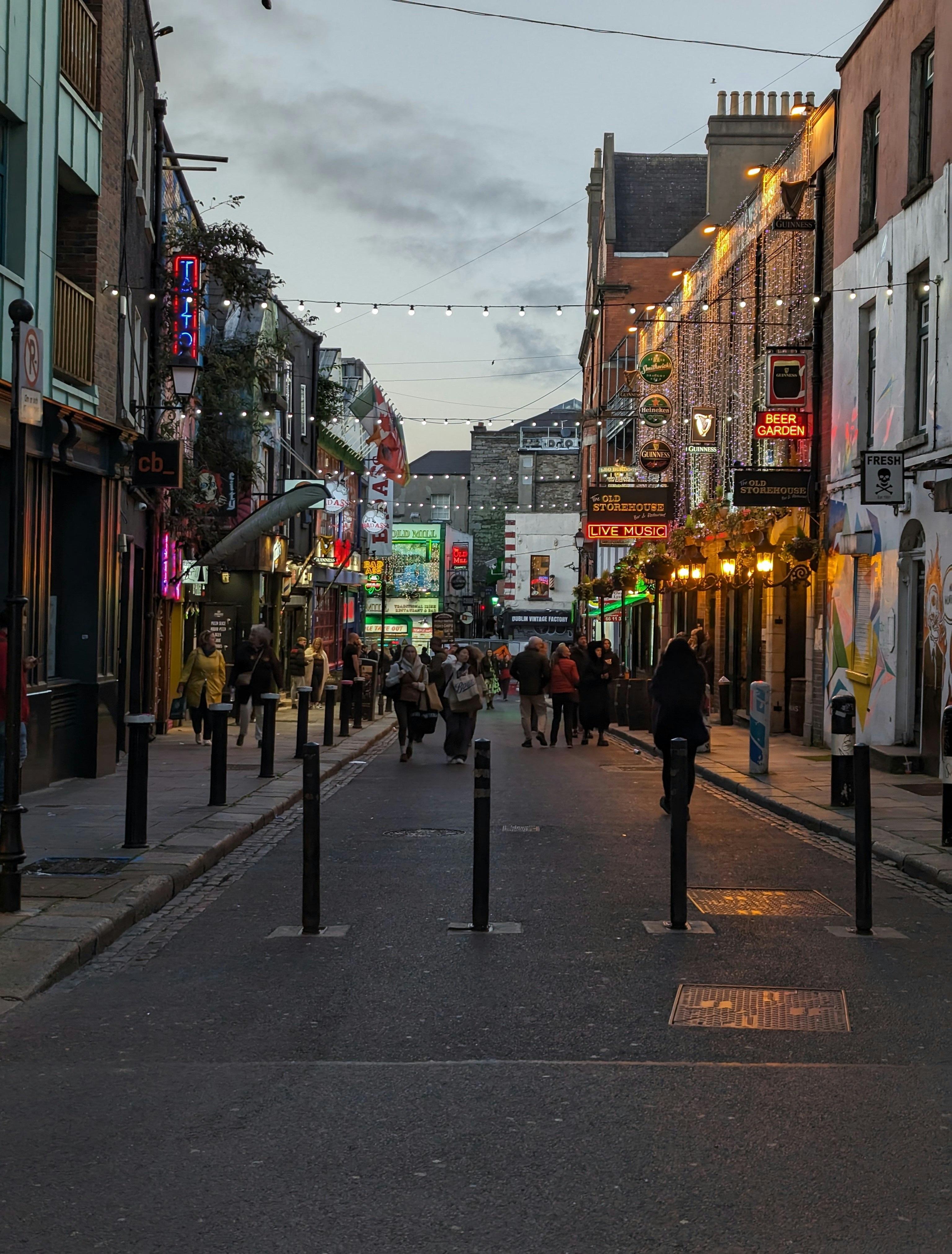 Photograph of a bustling urban street at dusk, lined with neon signs and string lights, as pedestrians walk between lit storefronts.
