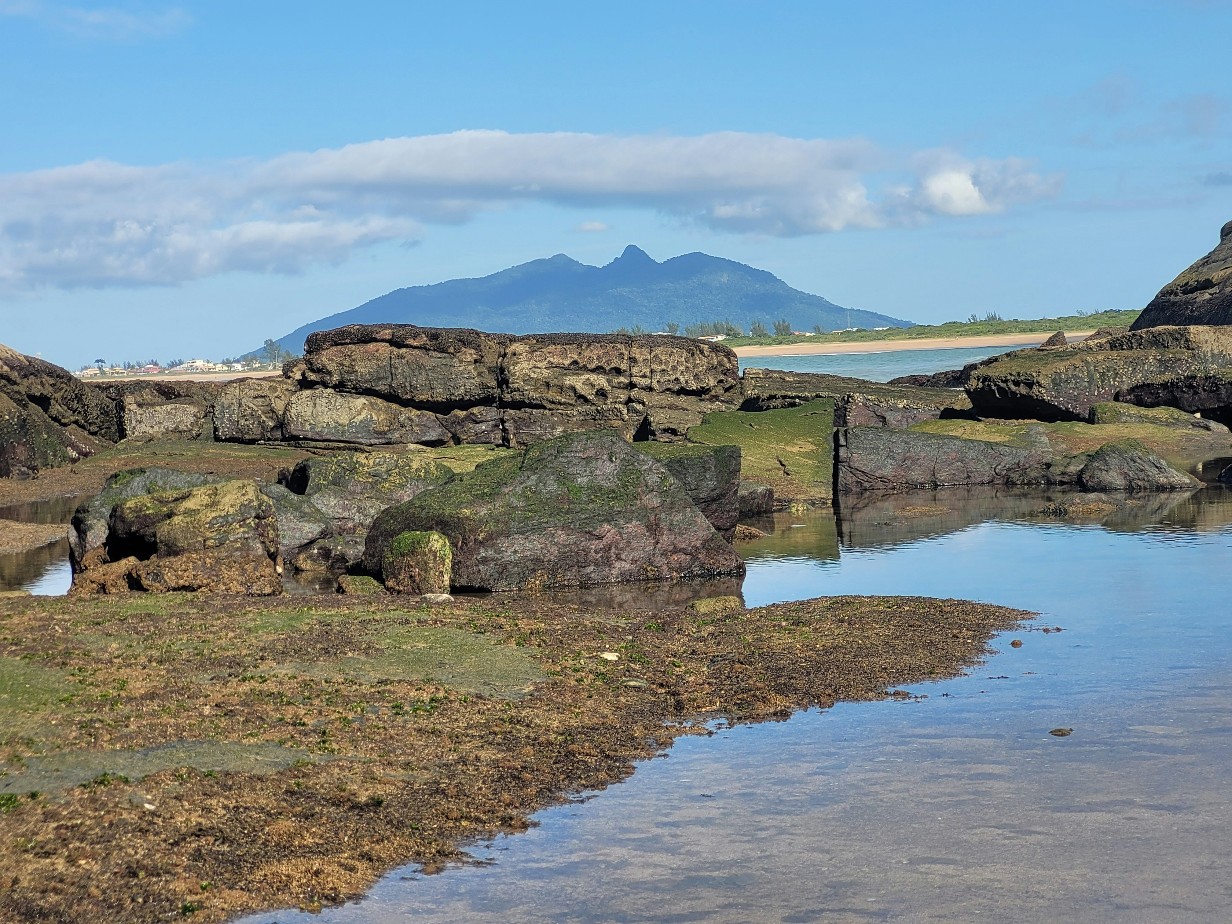 Tidal pools reflect rocky formations with a mountain range in the distant background under a clear blue sky.