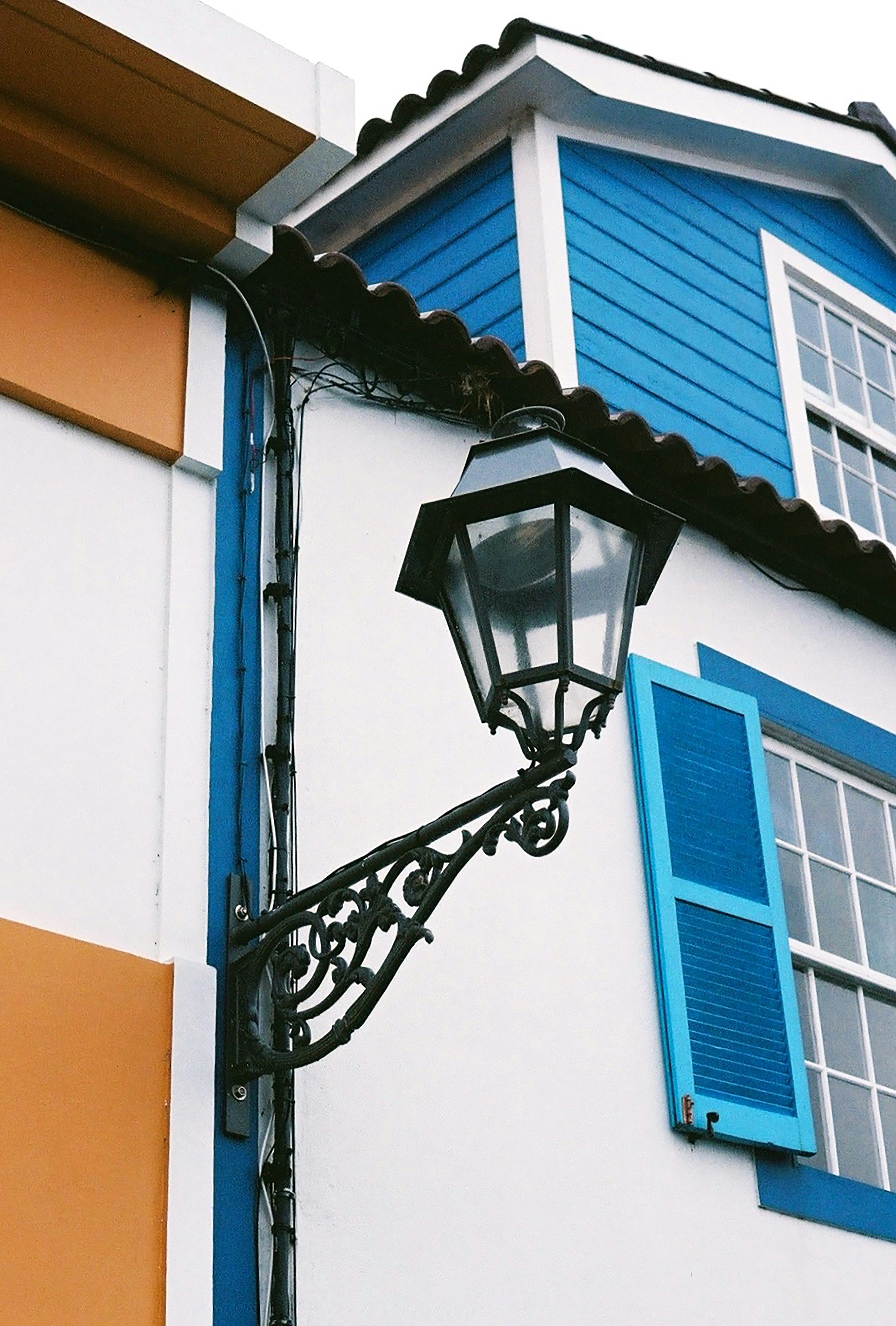 Vintage wrought-iron street lantern extends from a white wall framed by cobalt blue shutters and warm orange trim.