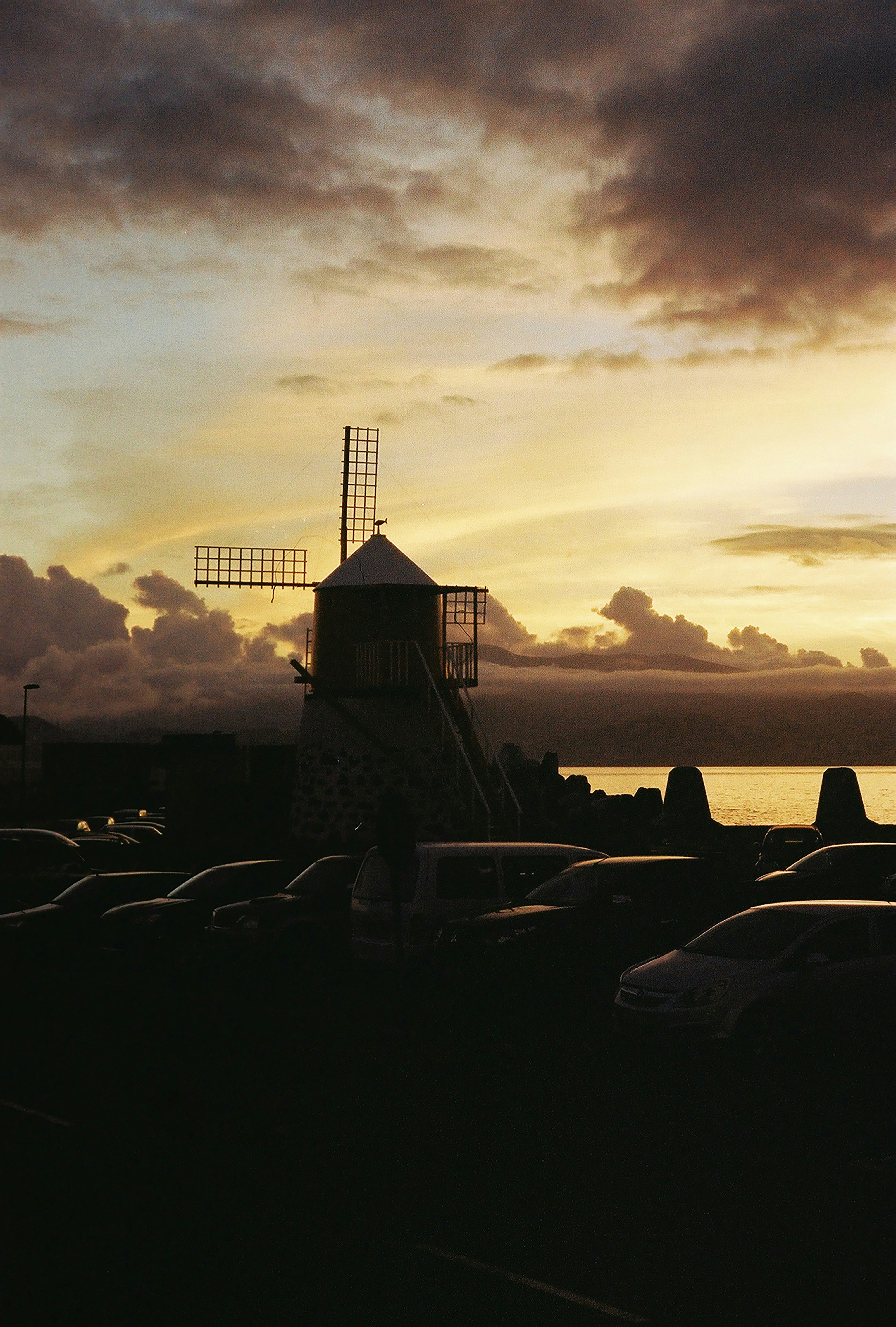 Windmill silhouette along a harbor at sunset, with parked cars in the foreground creating a moody, silhouetted scene.