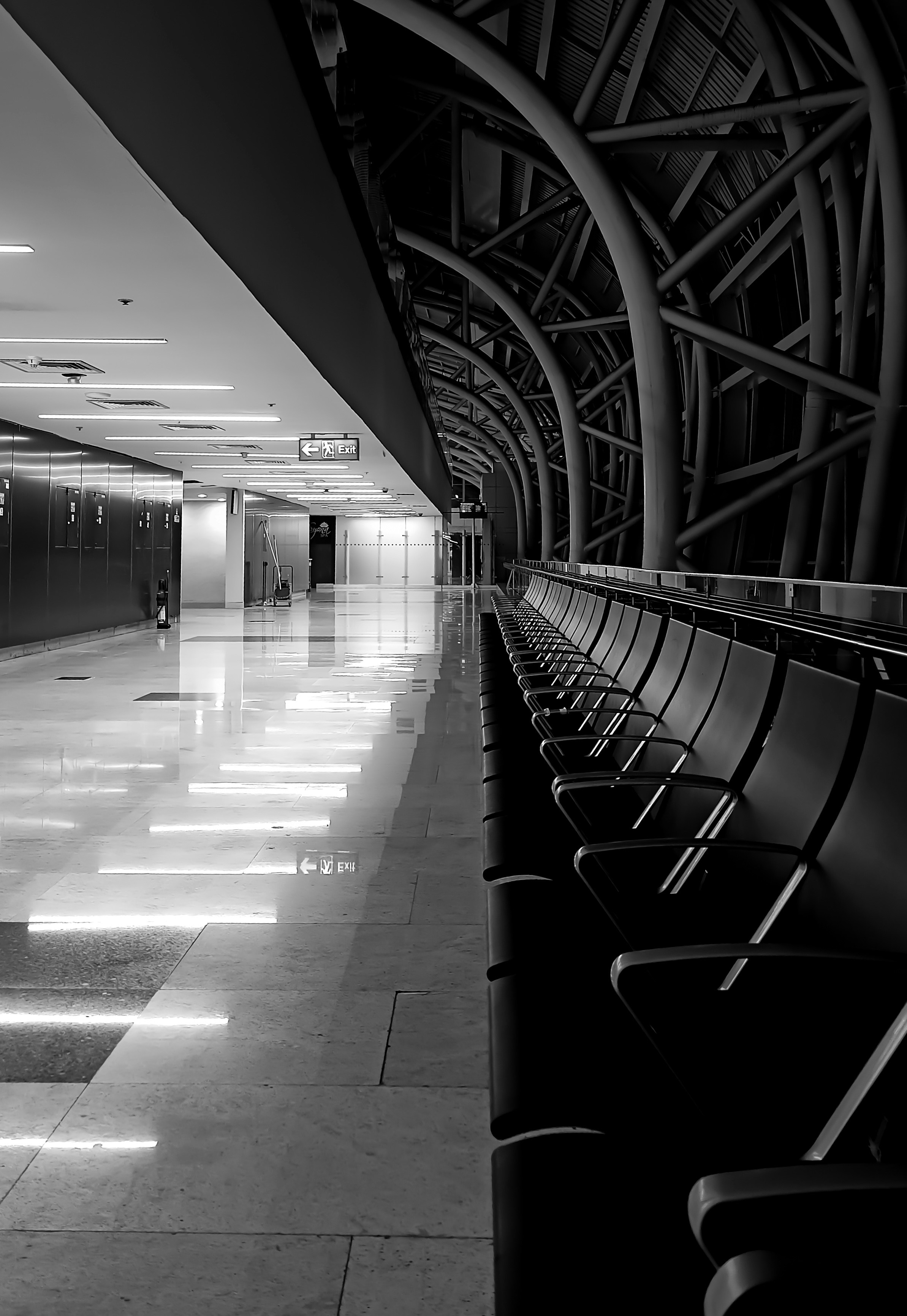 Long, reflective corridor of a modern transit hall with sculpted steel arches and rows of seats fading toward a bright doorway. The grayscale scene emphasizes architectural rhythm and emptiness.