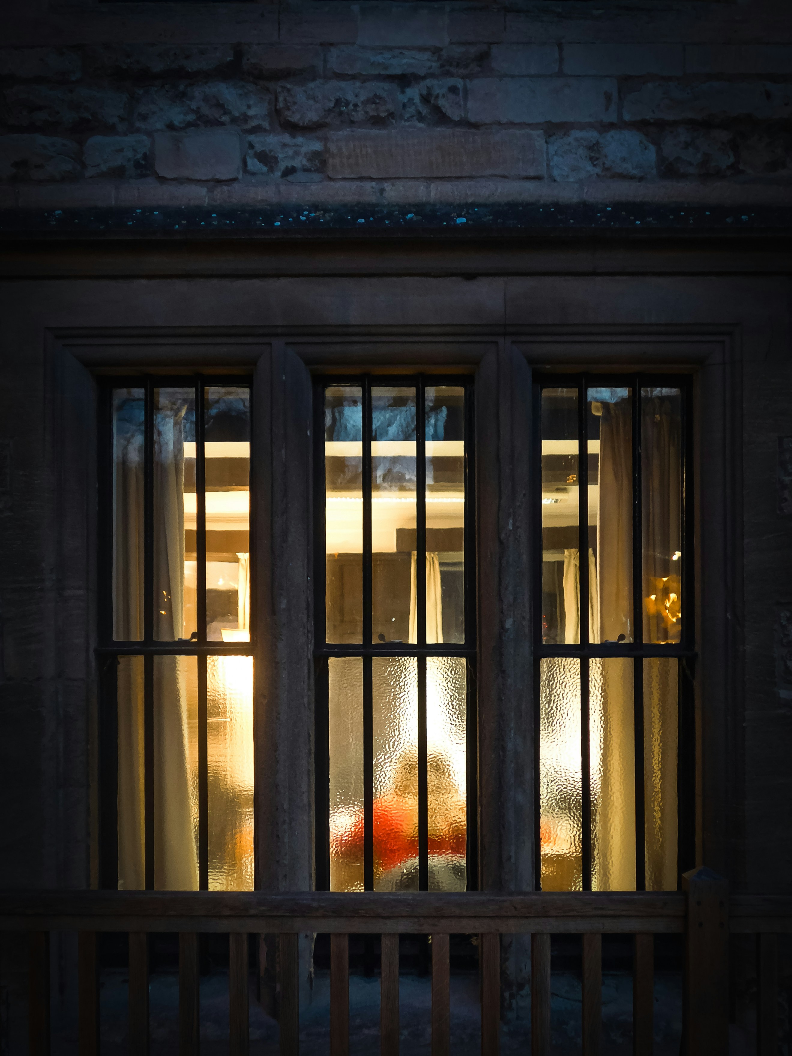 Soft interior light spills through a window with frosted glass and dark wooden frames on a weathered brick building.