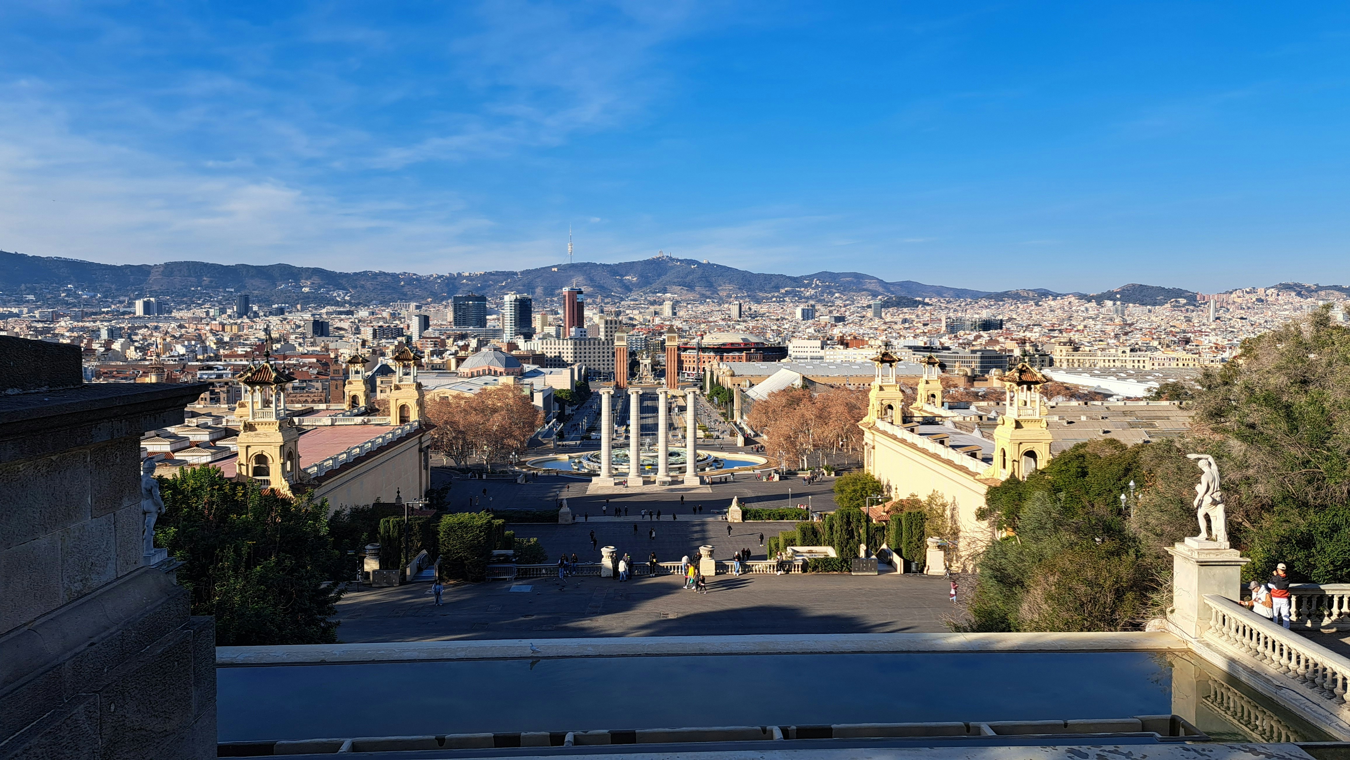 Wide cityscape view of Barcelona from Montjuïc, with a ceremonial staircase and statues in the foreground and a sunlit horizon of hills and rooftops under a clear blue sky.