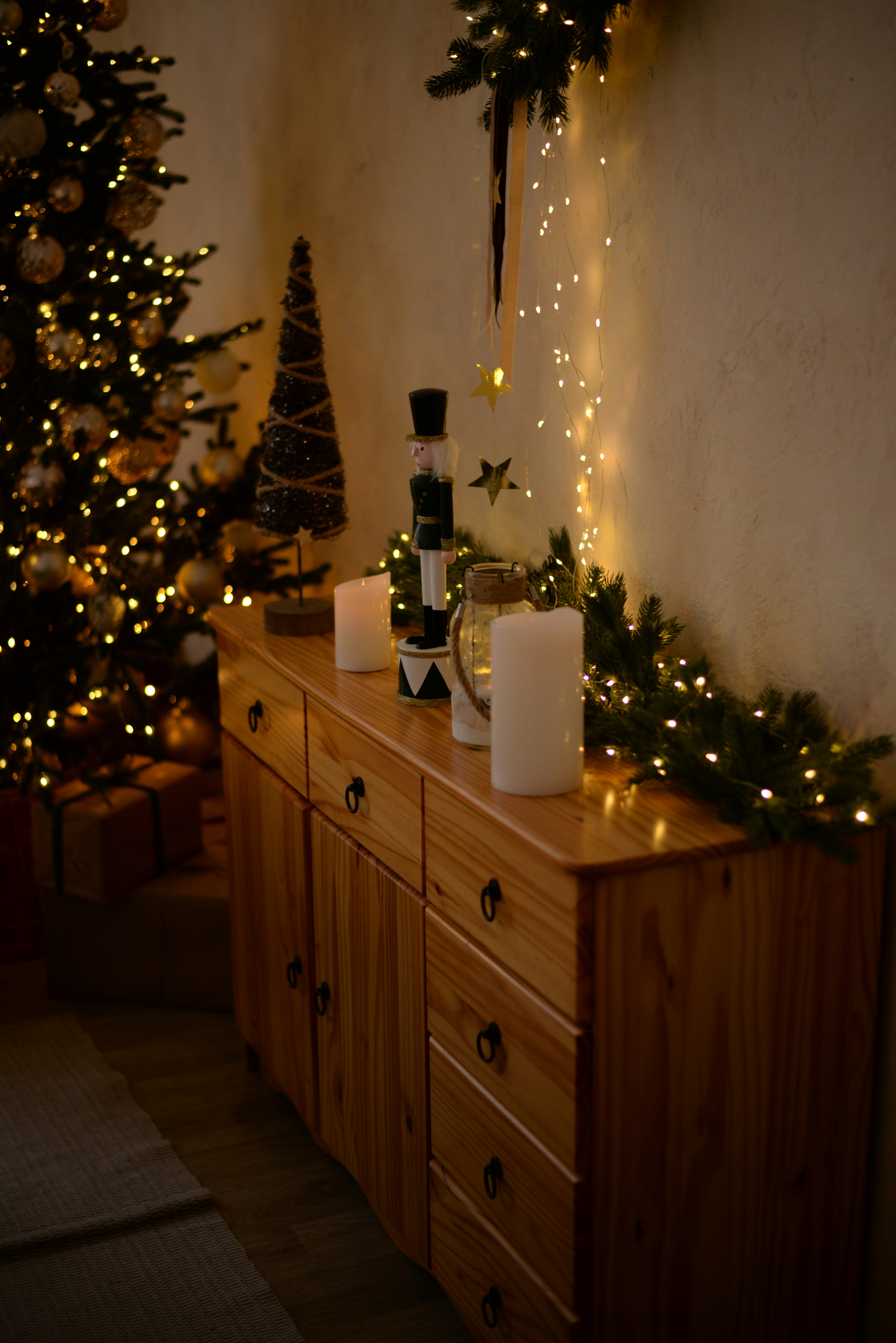 A warm Christmas vignette on a wooden sideboard with candles, garlands, and twinkling tree lights.
