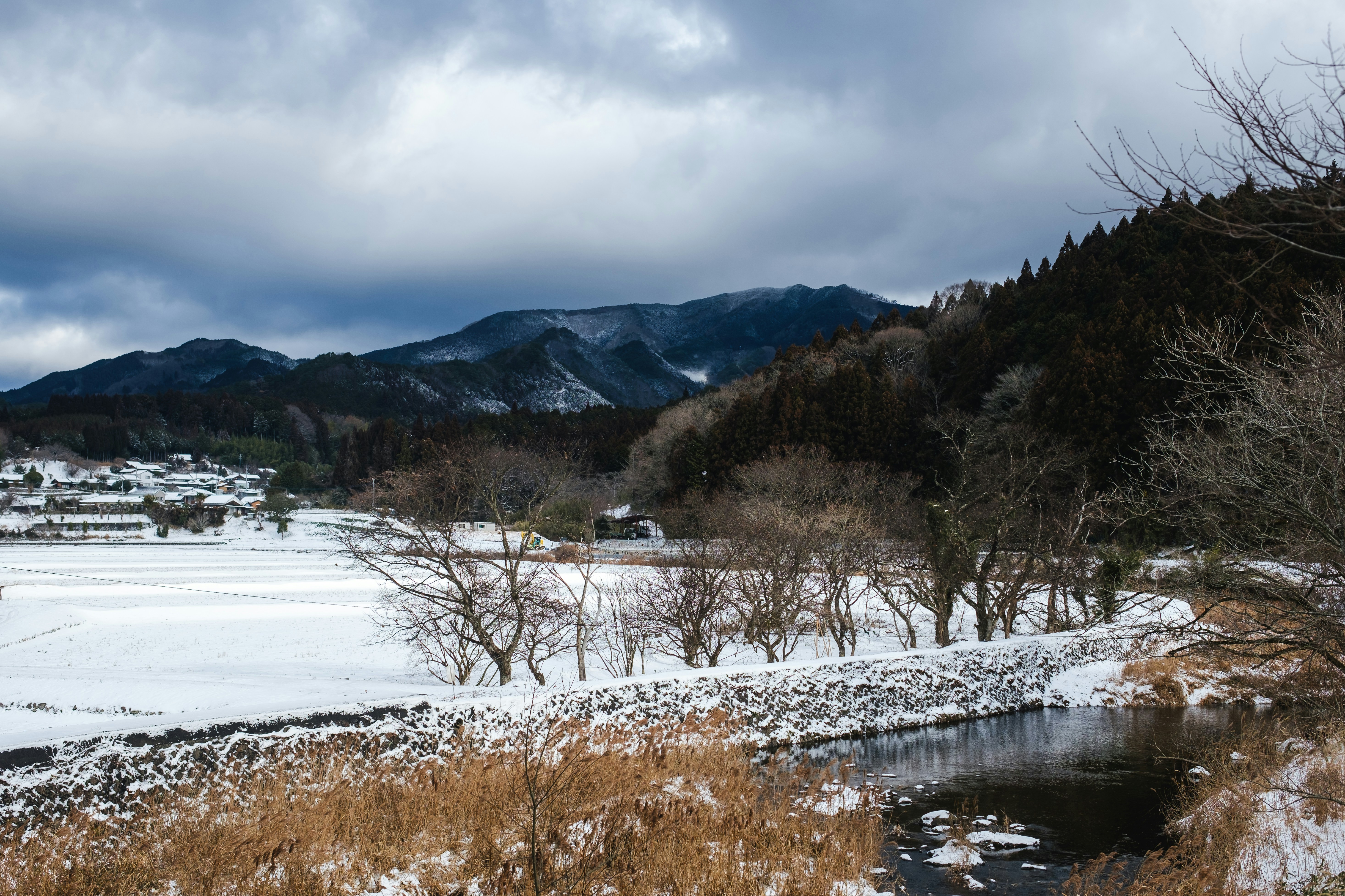 Snow-covered fields stretch beneath a dramatic sky, framed by bare trees and distant mountains. A gentle stream weaves through the scene.