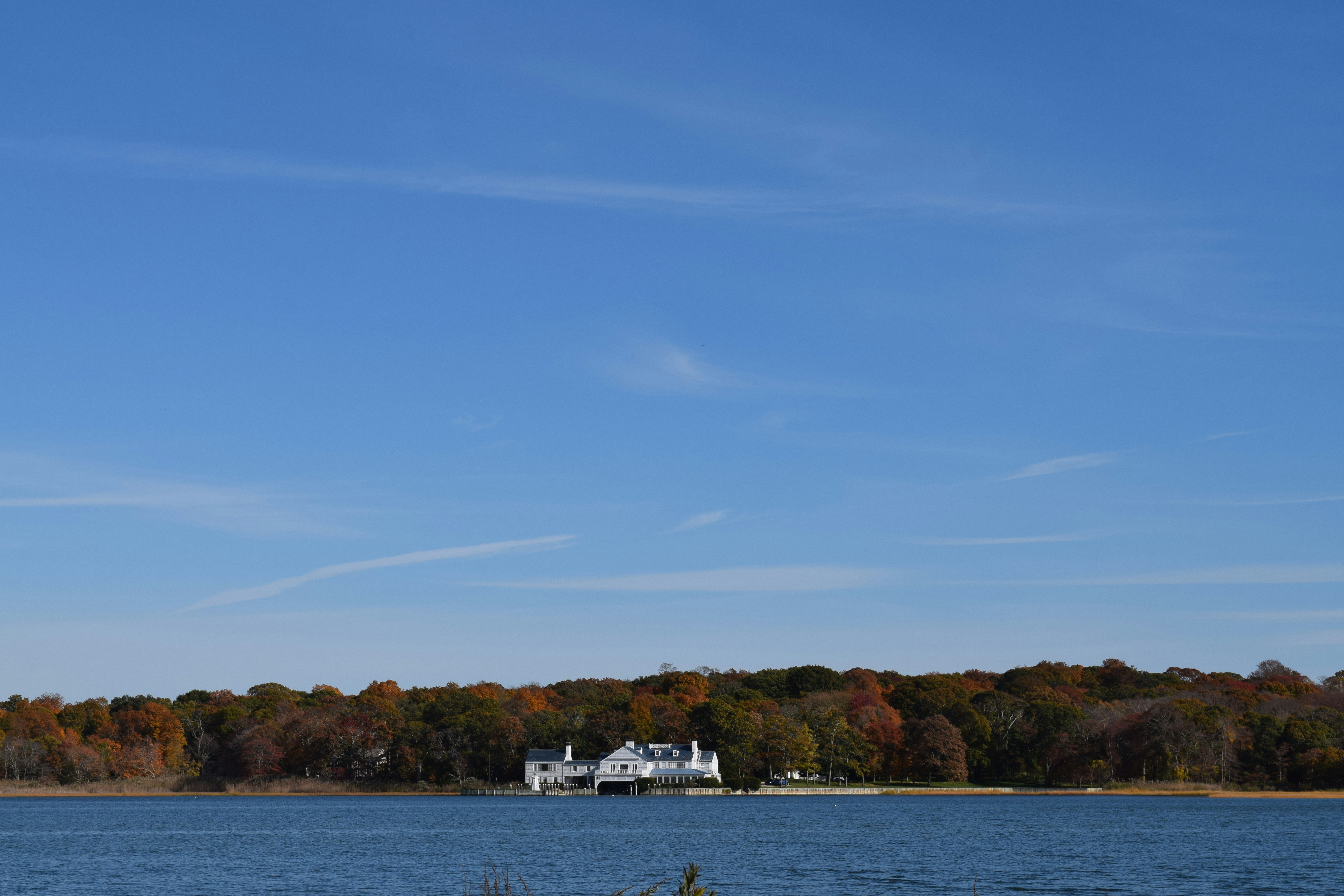 White house nestled by a lake with a backdrop of autumn foliage under a clear blue sky.