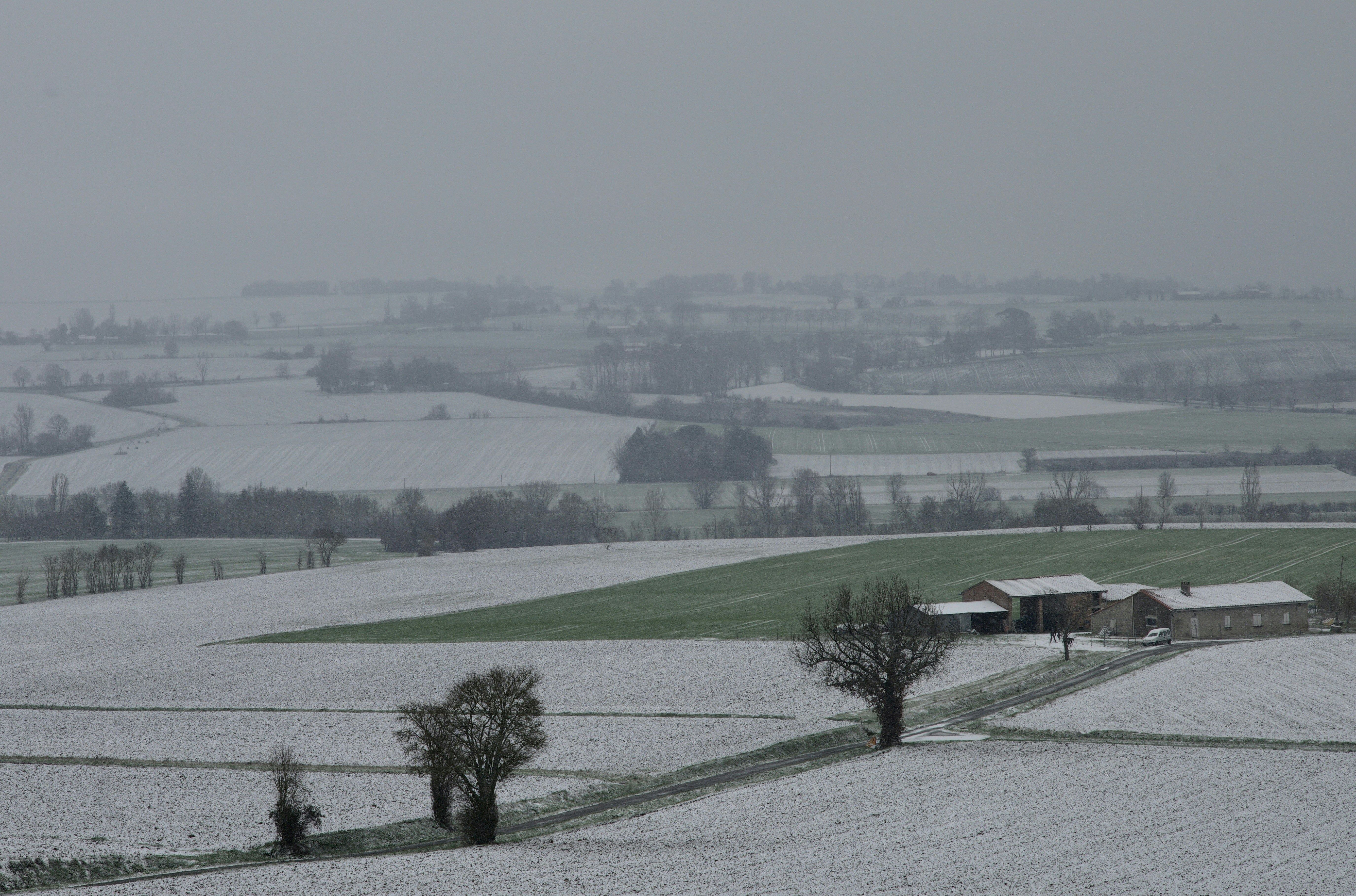 Snow-dusted fields stretch to a distant horizon with a small farmstead and scattered trees under an overcast winter sky.