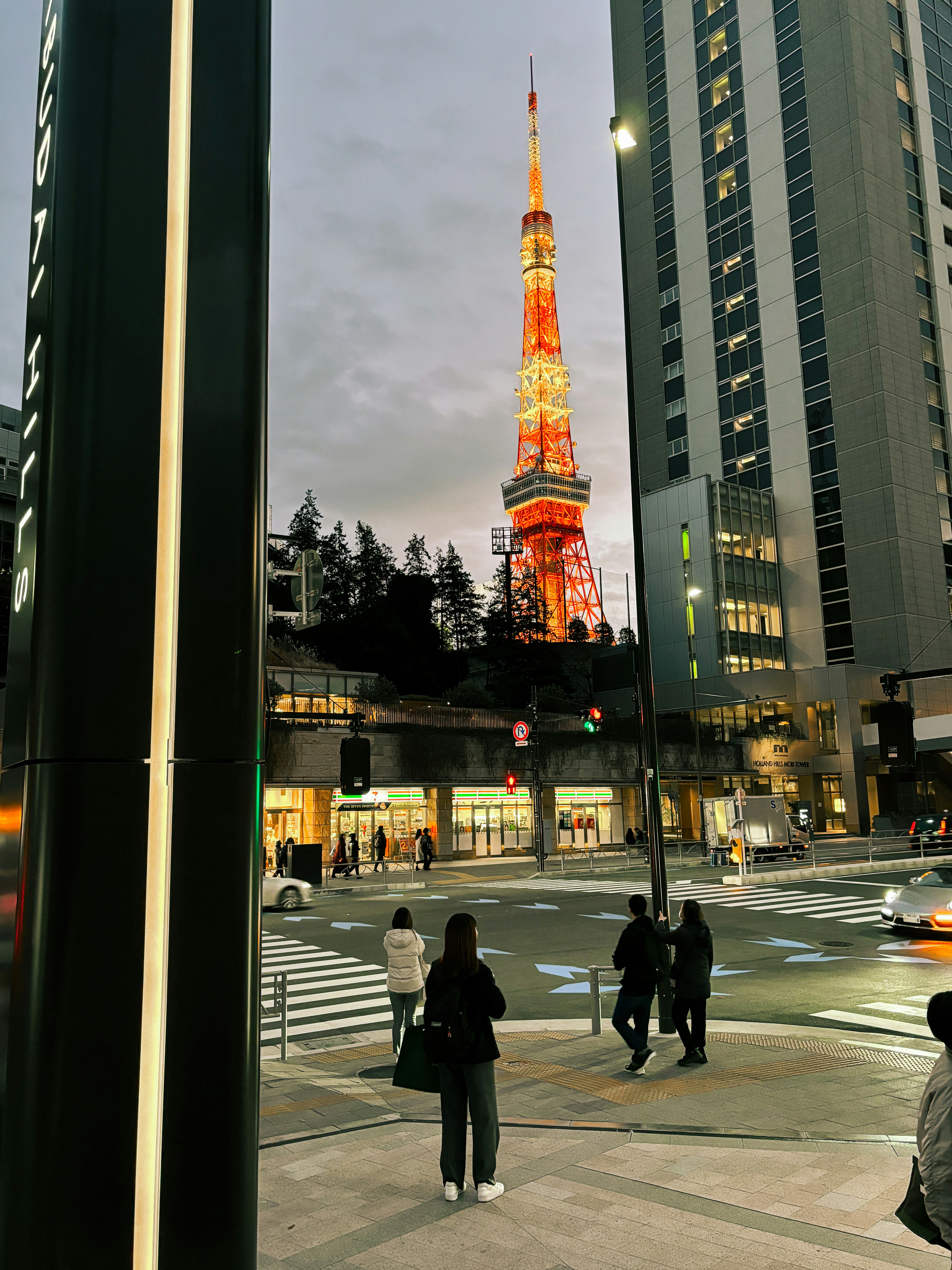 Photograph of Tokyo Tower lit in orange behind a busy city street with pedestrians crossing at dusk.