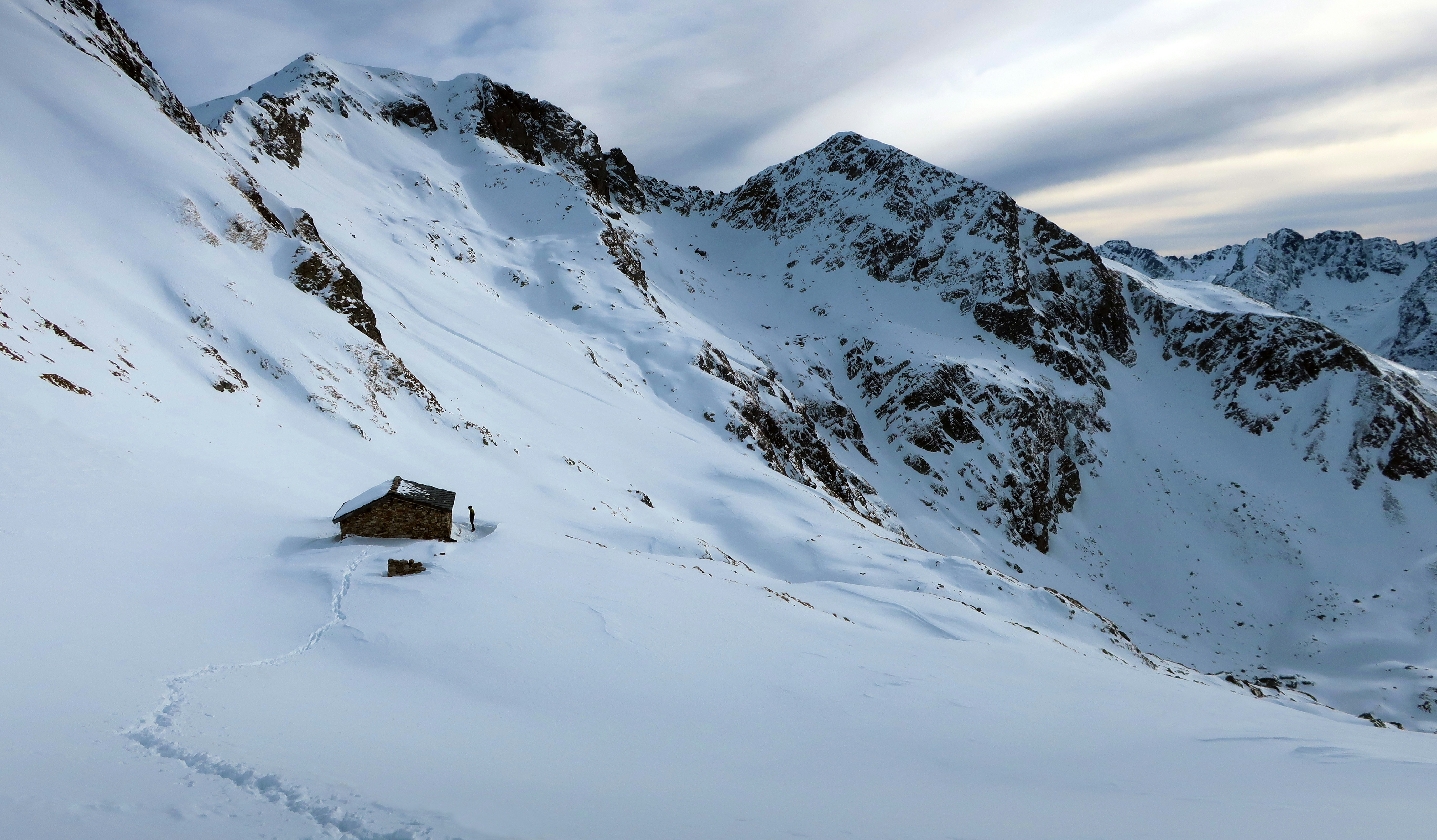 A rustic cabin nestled in a vast snowy landscape, surrounded by towering mountains under a cloudy sky.