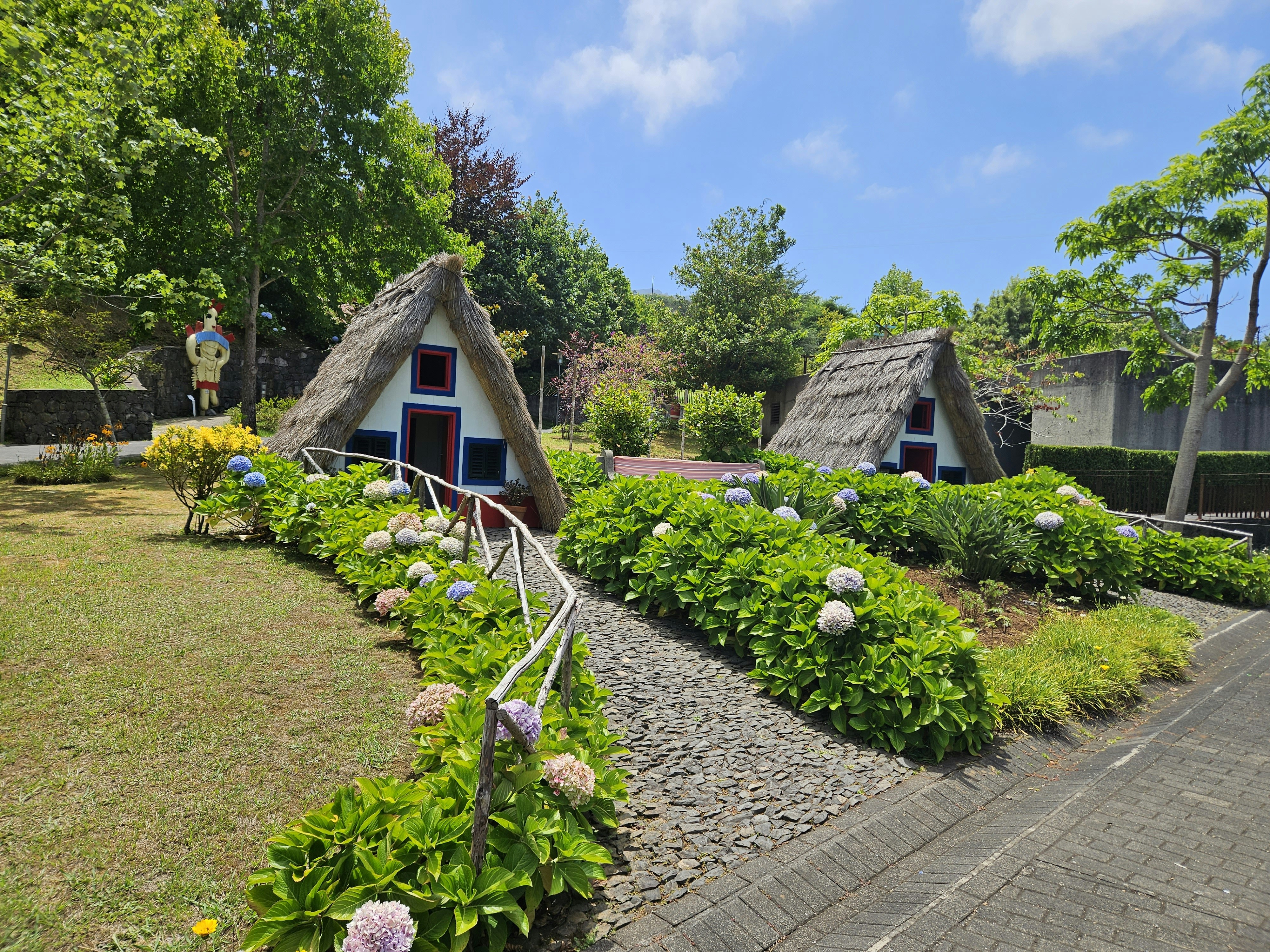 Sunlit garden featuring two thatched-roof cottages flanked by hydrangea bushes along a cobblestone path.