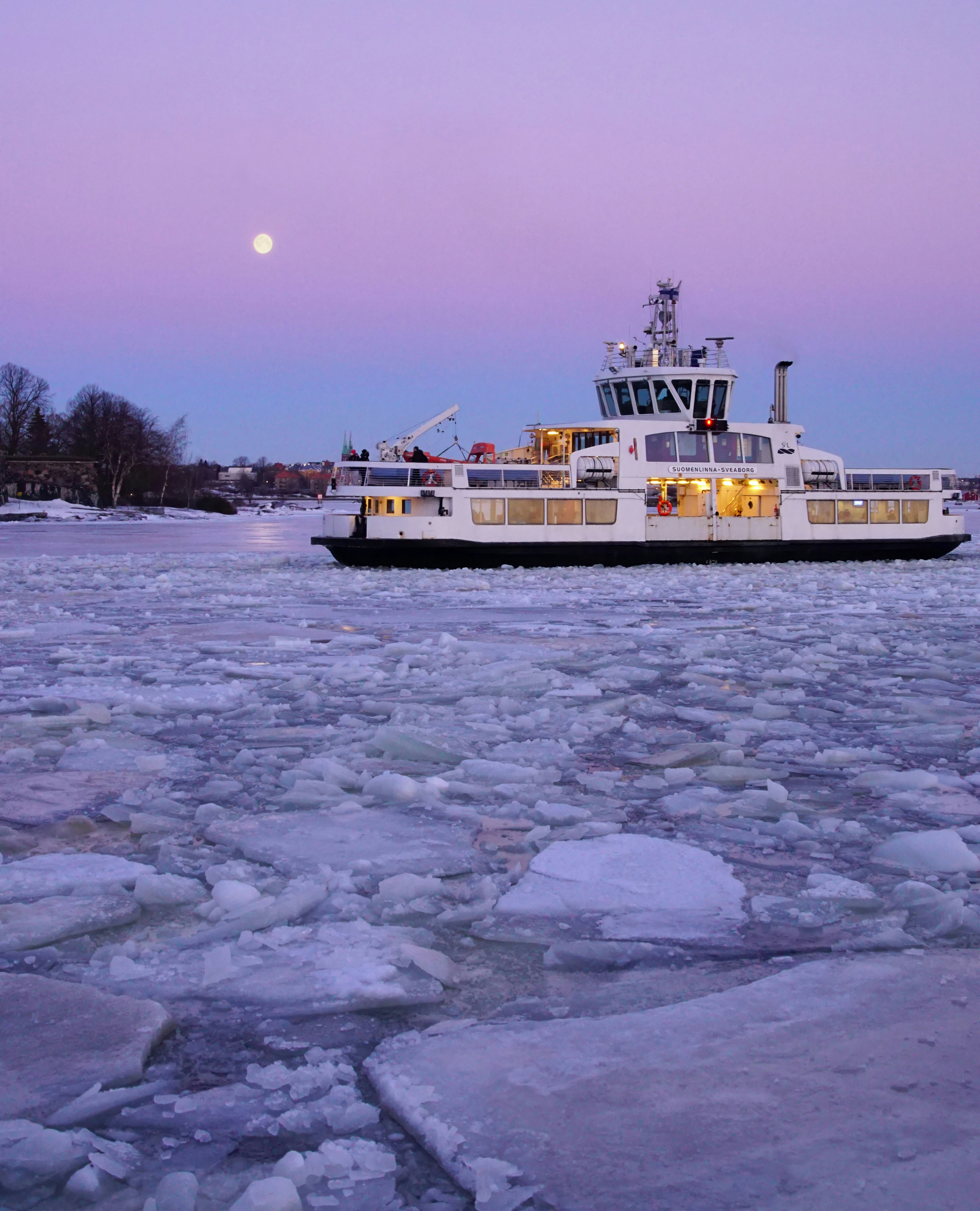 A ferry pushing through icy waters in Suomenlinna, Finland in the early morning with the moon setting in the background. | a large boat floating on top of ice covered water