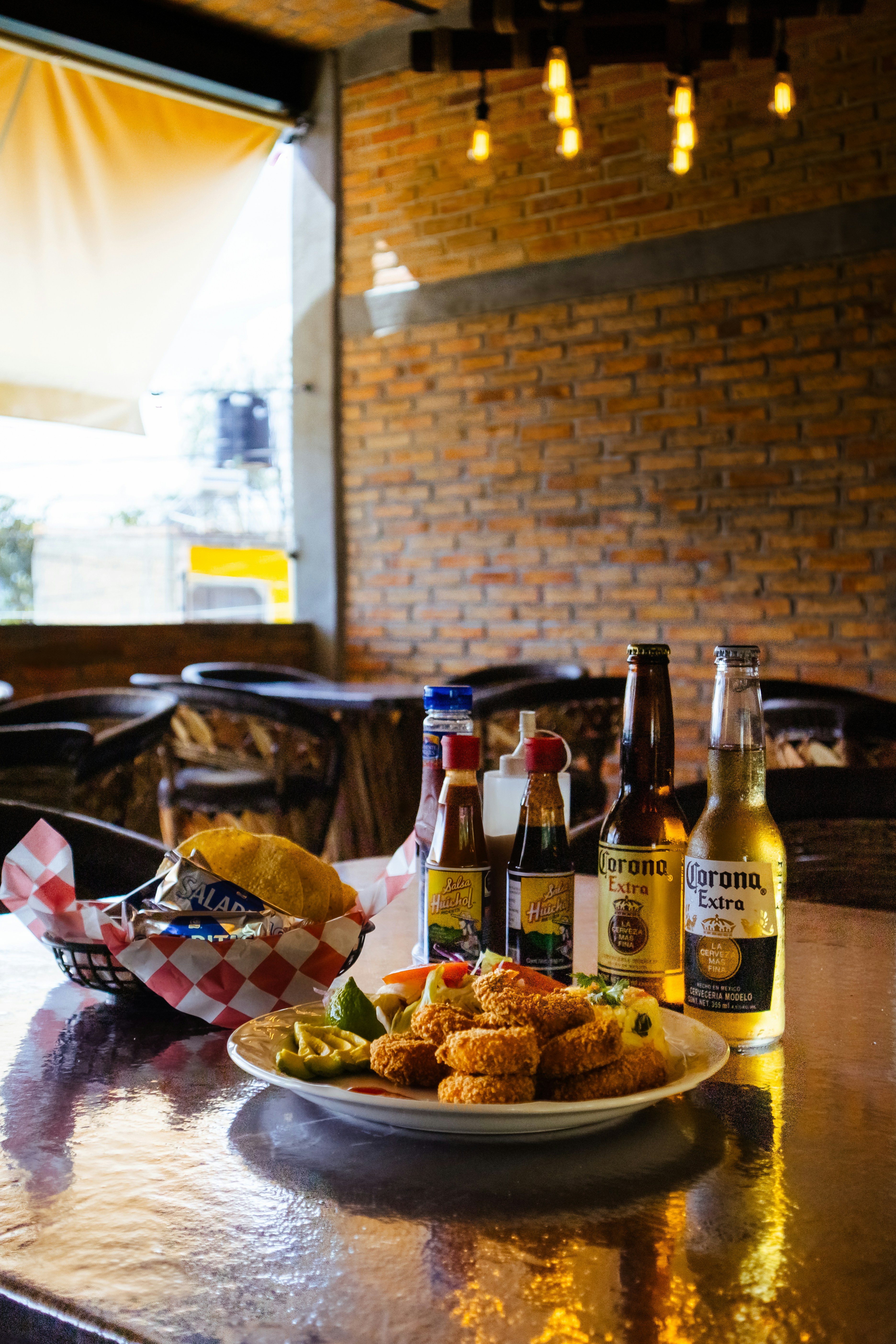 Casual restaurant photo showing a plate of crispy fried bites with lime, a basket of fries, and sauces. Corona bottles stand beside the plate on a reflective table, set against a brick wall and warm pendant lighting.
