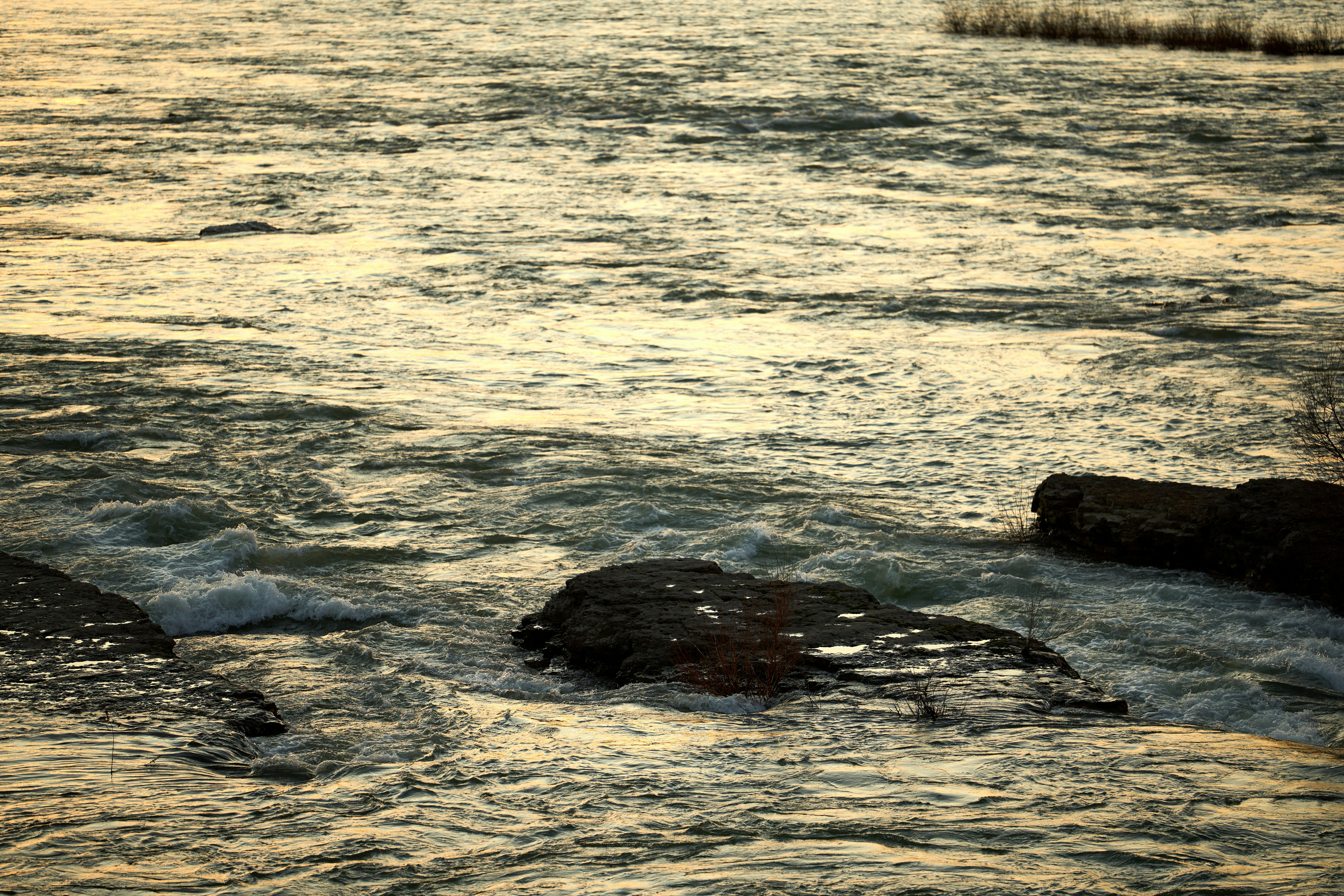 Gentle waves caress the rugged rocks as the river reflects the warm hues of the setting sun.