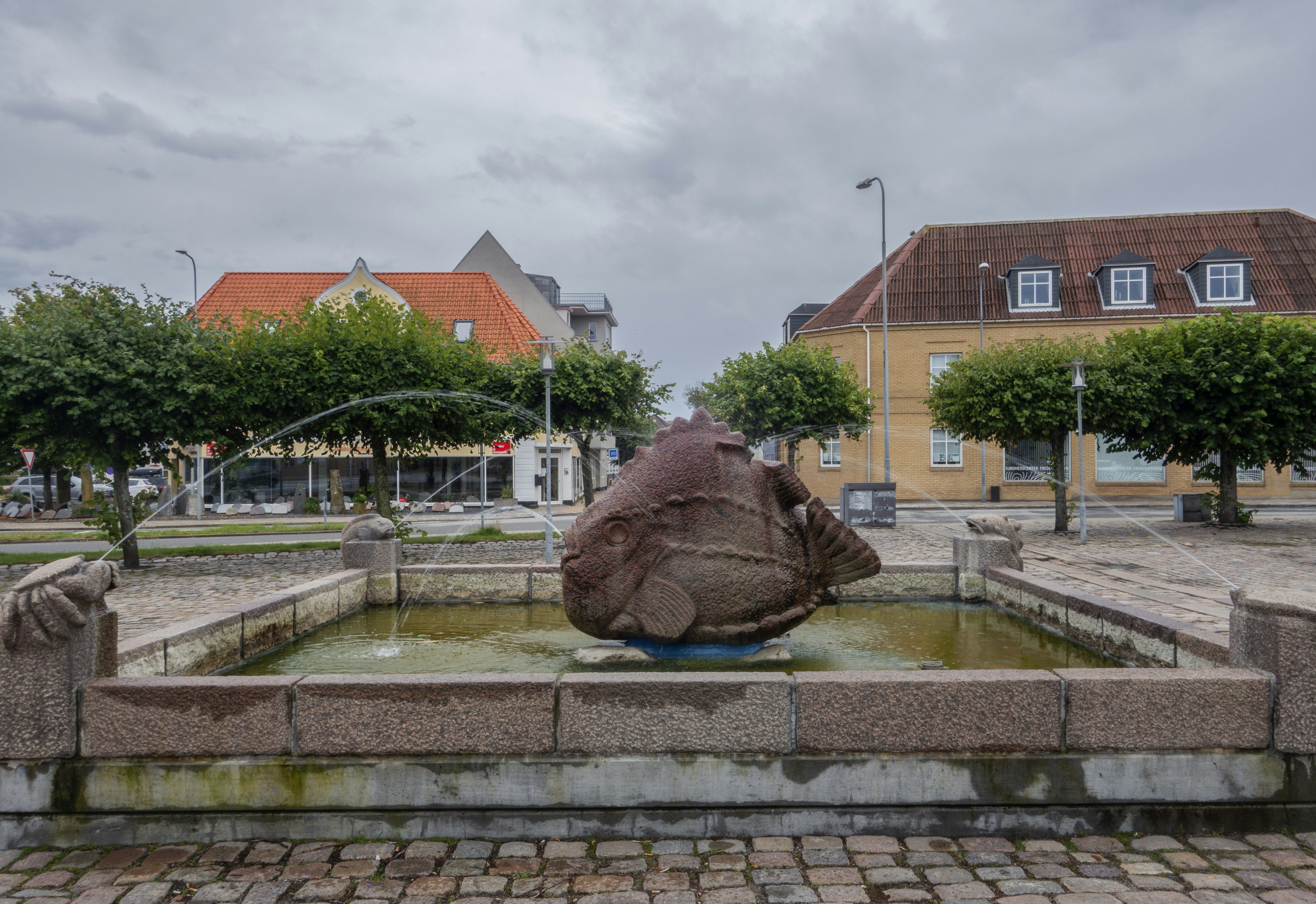 Bronze sculpture in a quaint Danish town square surrounded by trimmed trees and historic buildings under a cloudy sky.