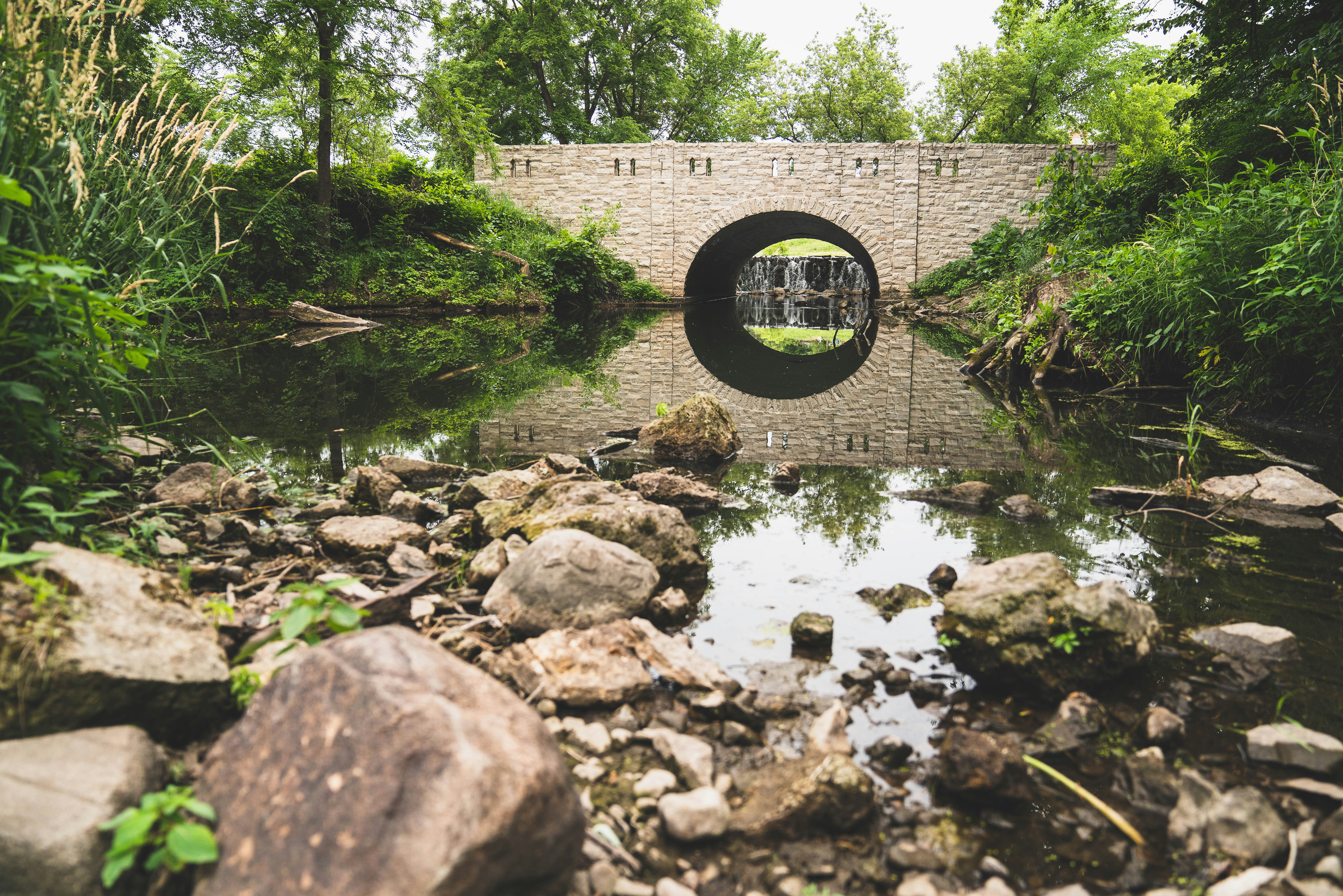 Stone bridge arches over a tranquil stream surrounded by lush greenery.