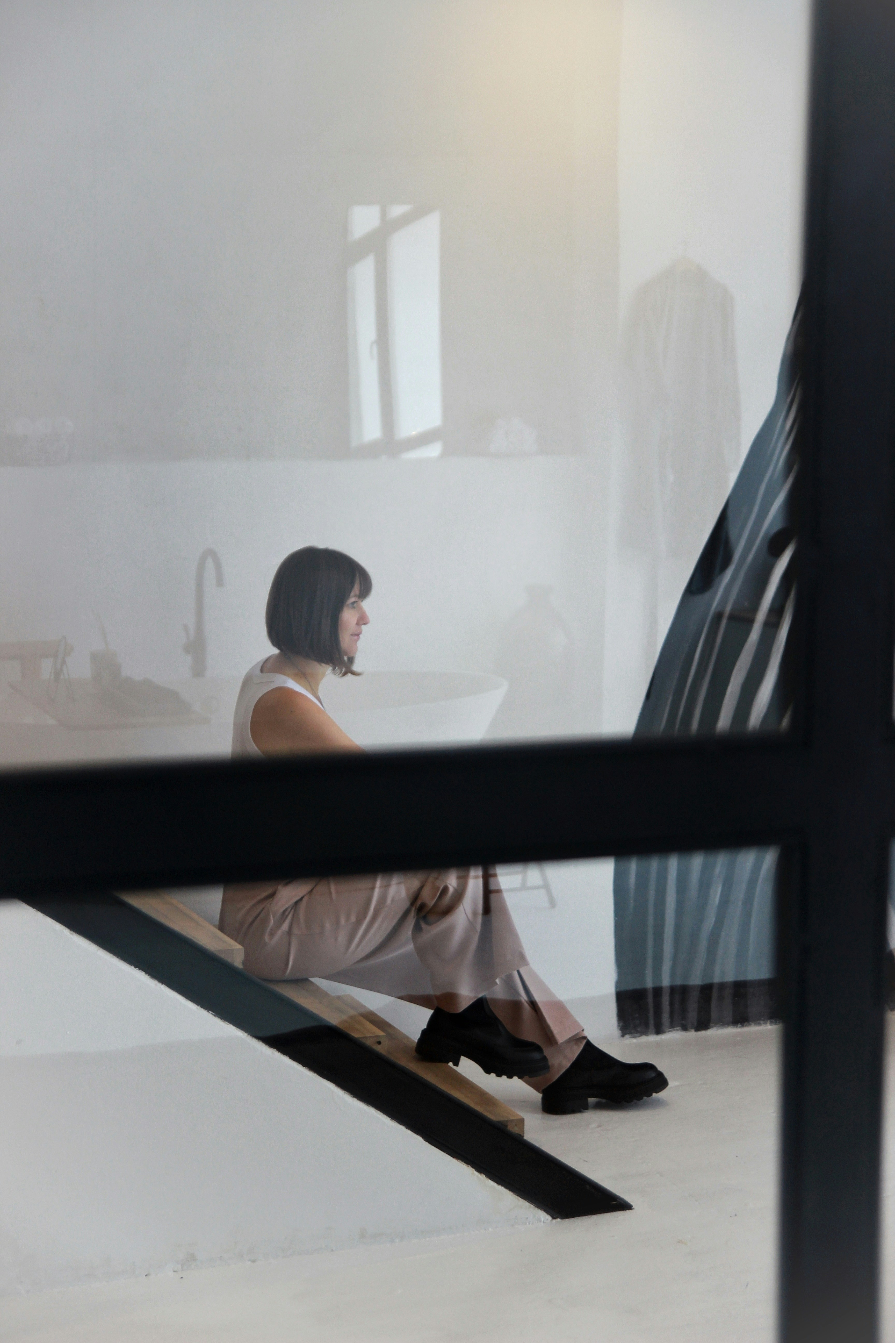 A woman seated in a minimalist room, partially obscured by a glass partition, with soft natural light illuminating her figure.