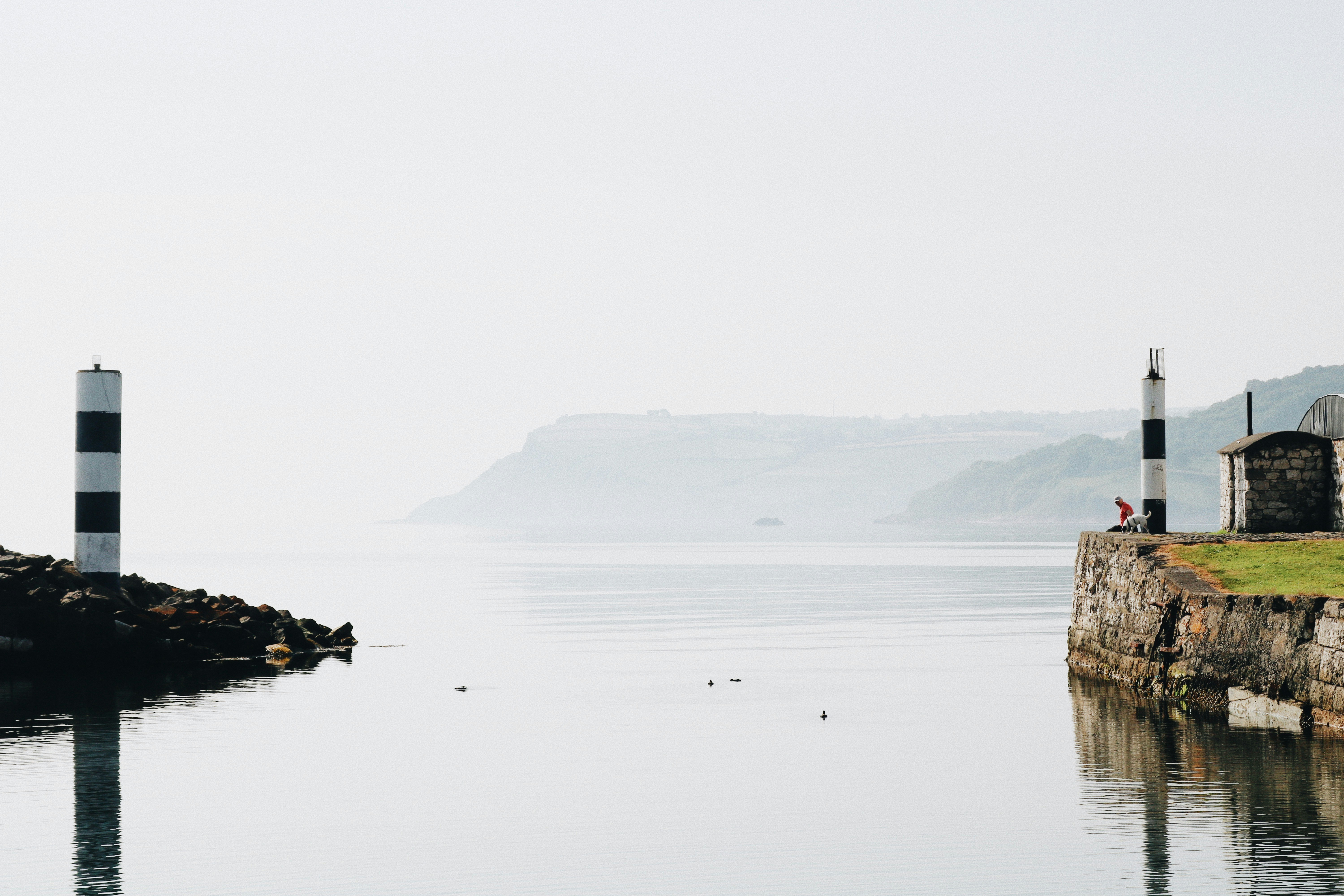 Two striped lighthouse markers stand sentinel by a calm harbor, with a solitary figure engaged in contemplation on the stone pier.