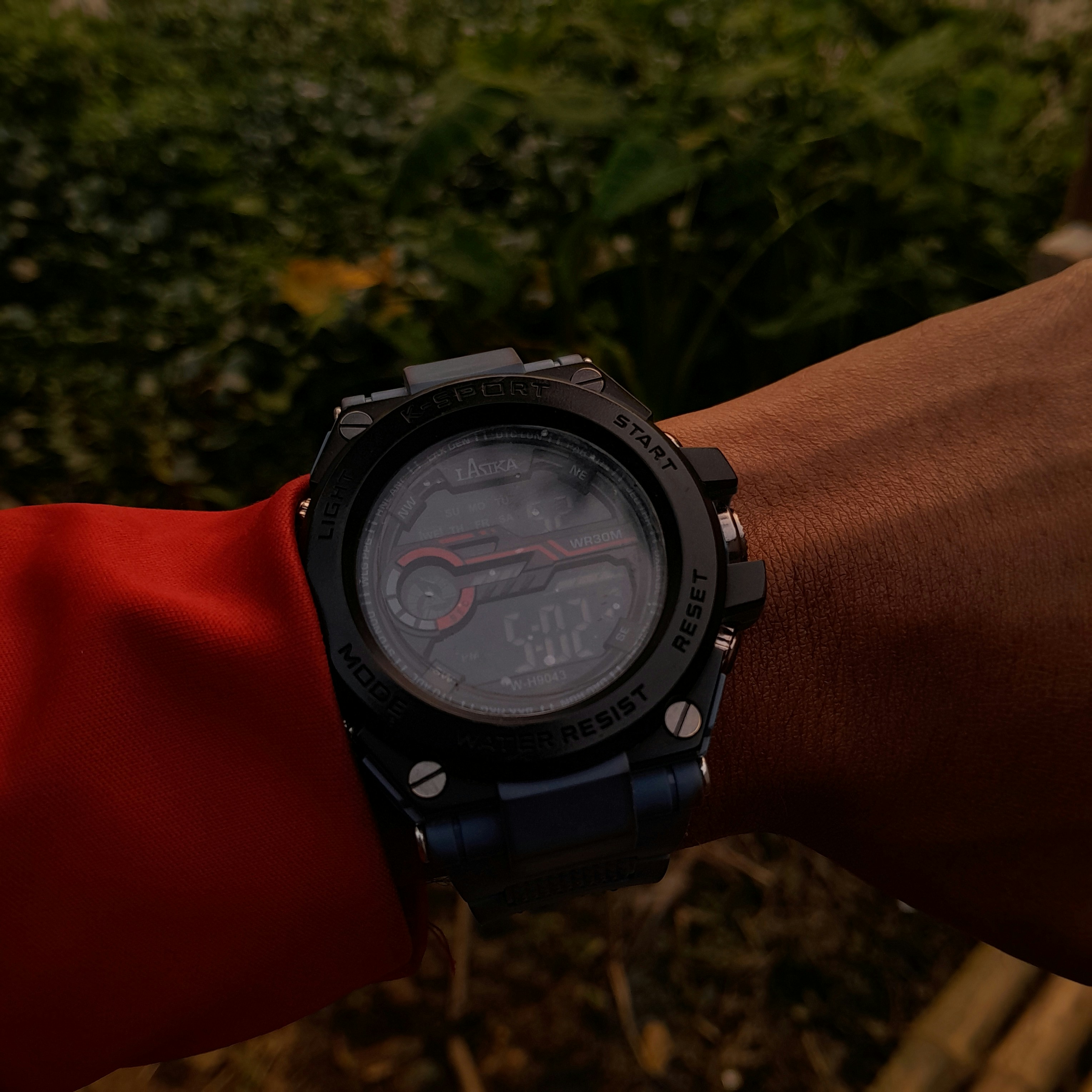 Close-up photograph of a rugged digital wristwatch on a red-sleeved arm, set against dark outdoor foliage.