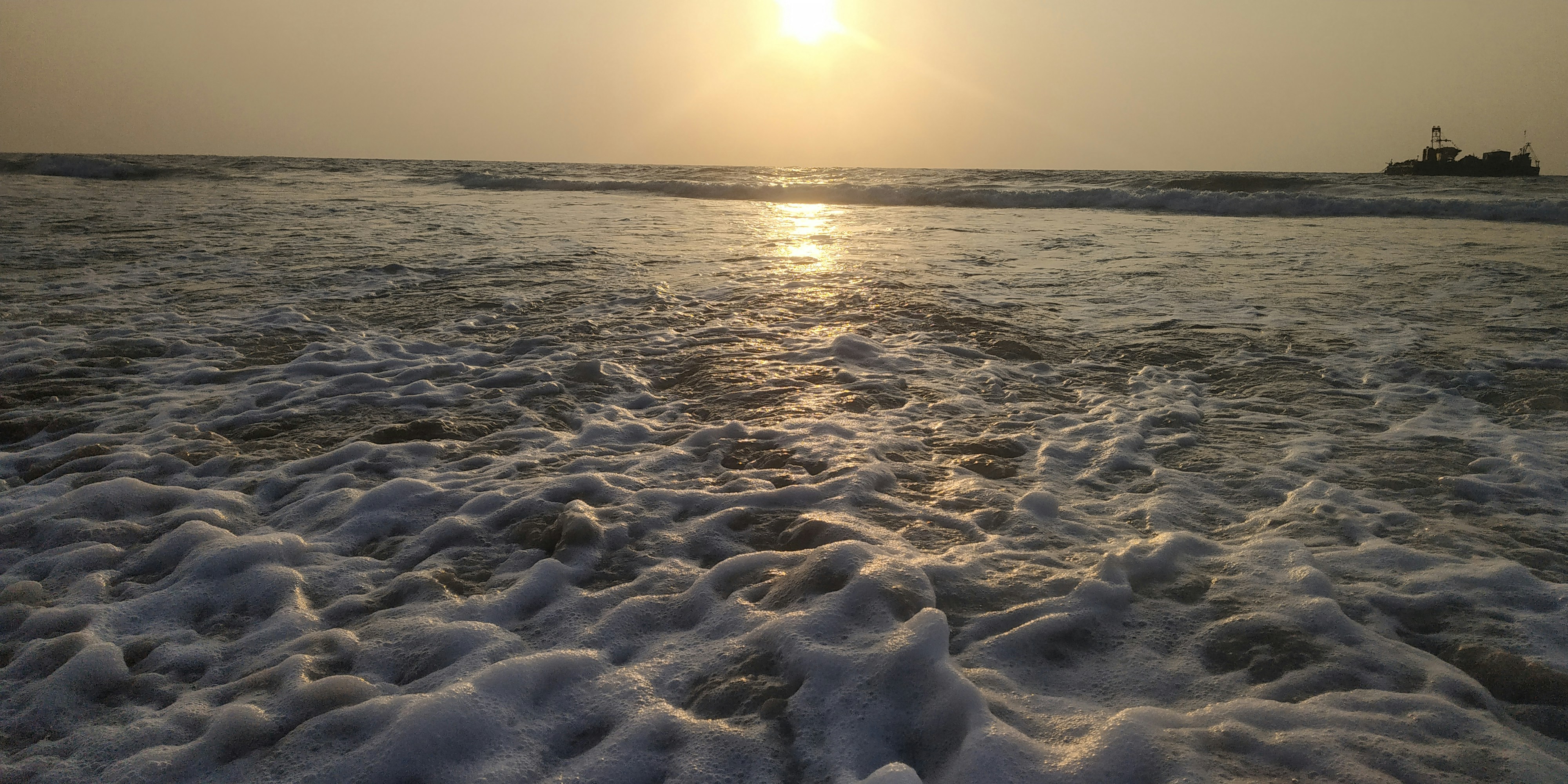 Foamy ocean waves under a warm, golden sunset with a distant ship on the horizon.