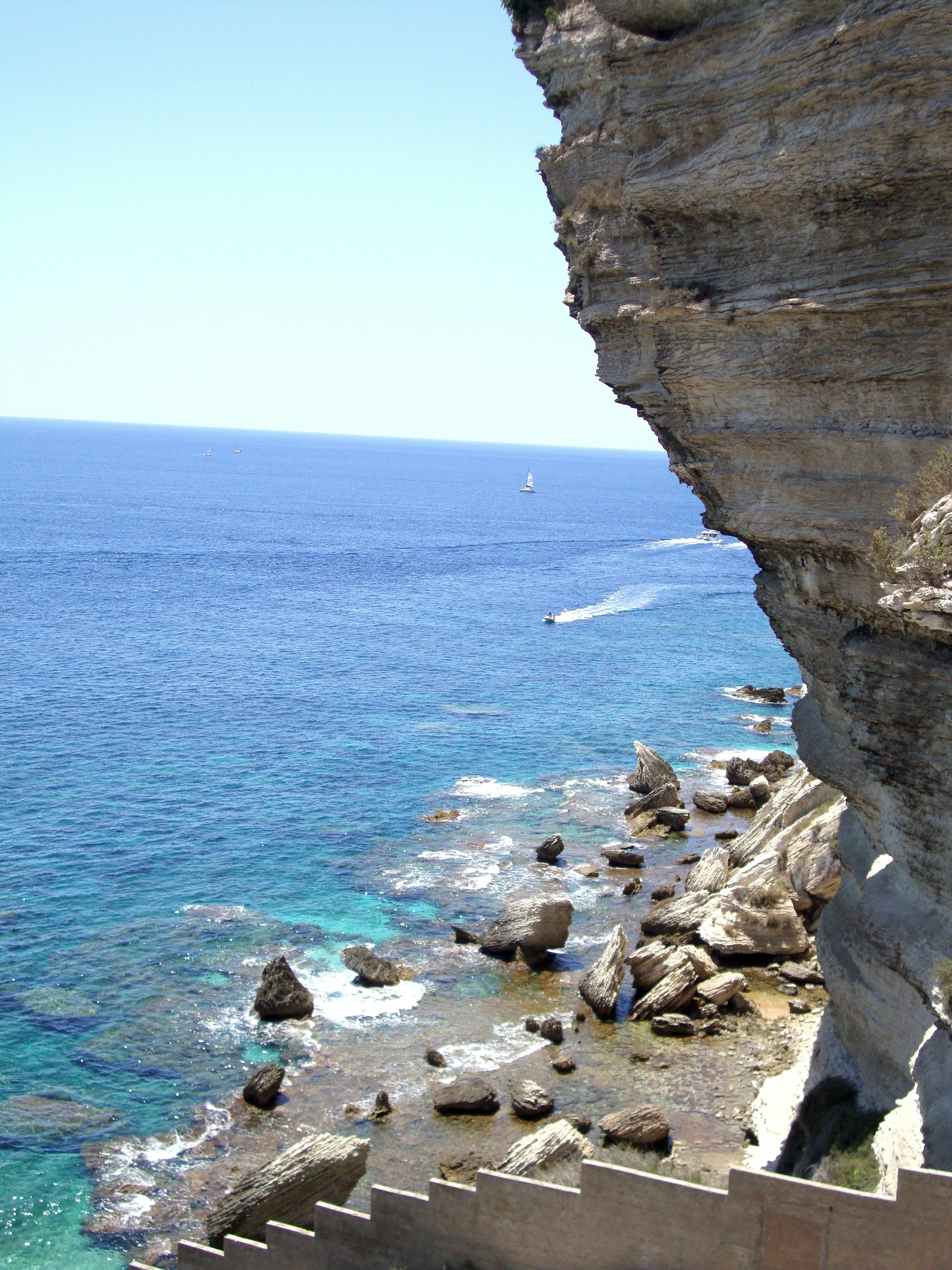 Sunlit limestone cliffs loom over a rocky shoreline, with a stairway inviting descent to the turquoise water. The scene captures a rugged coastal edge framed against a calm sea on a bright day.
