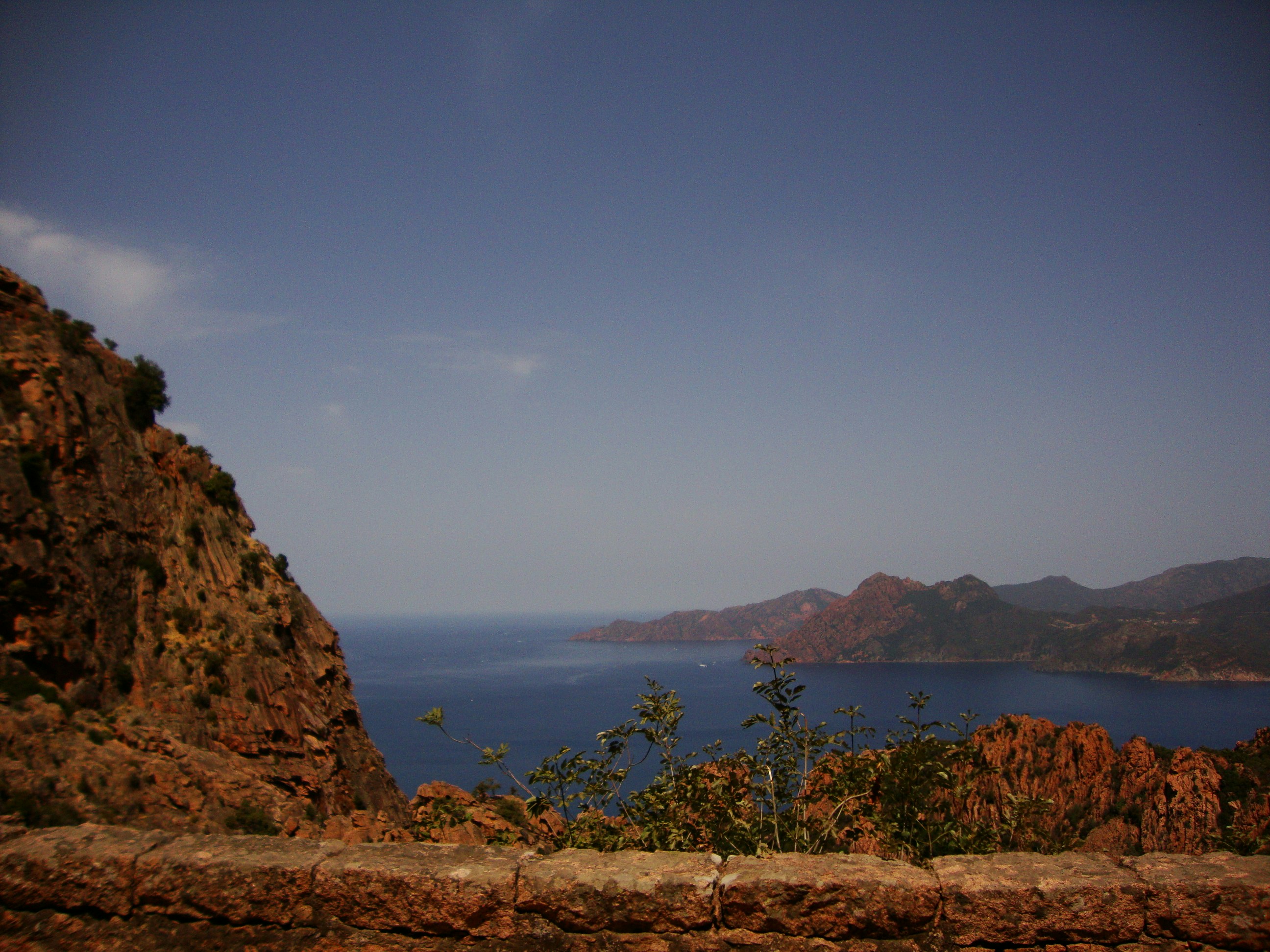 Rugged cliff edges frame a tranquil blue sea with distant headlands and islands under a clear sky.
