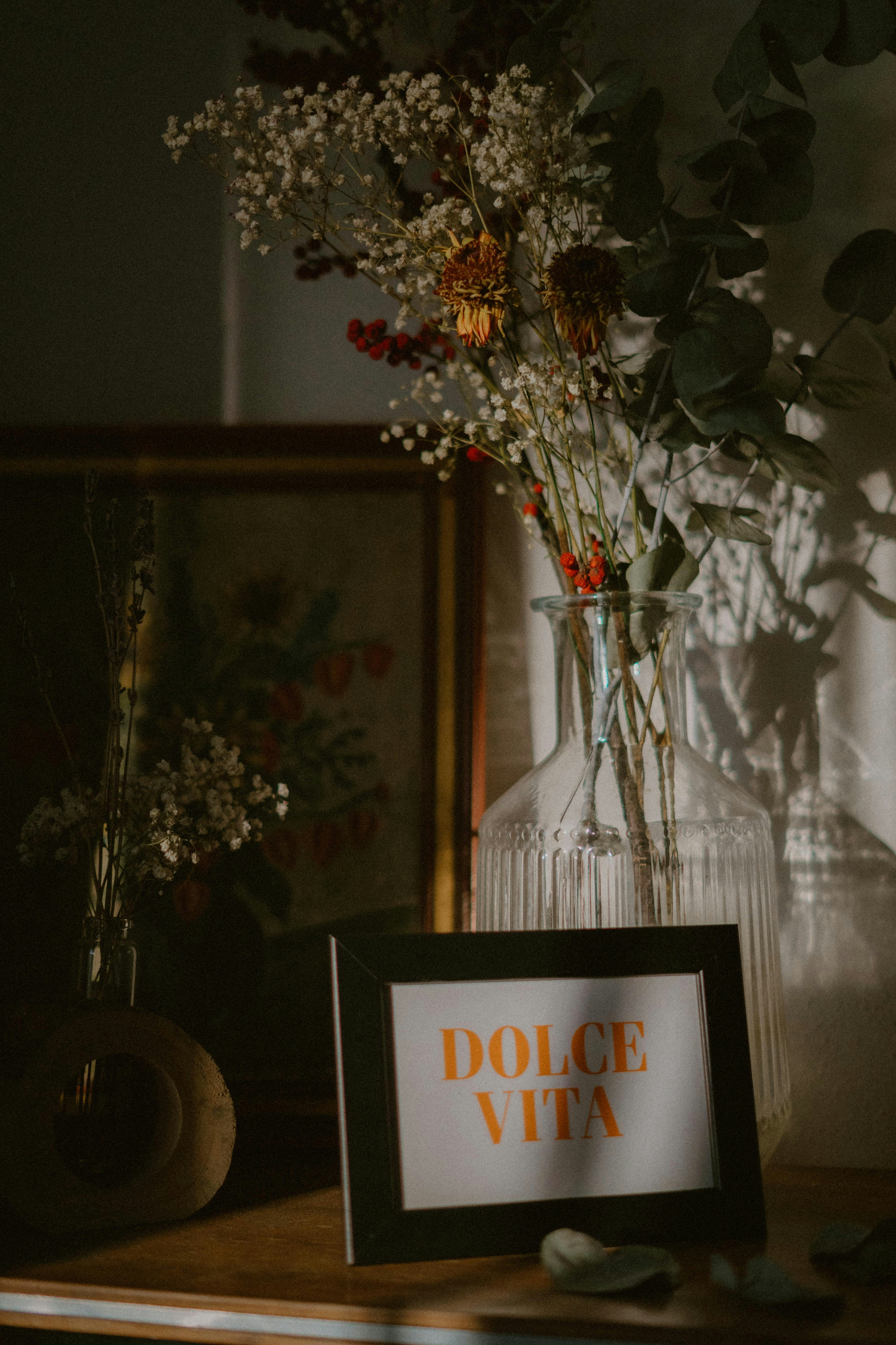 Dolce Vita in Dim Light: Dried Florals, Glass Vase, and a Framed Orange-Text Whisper