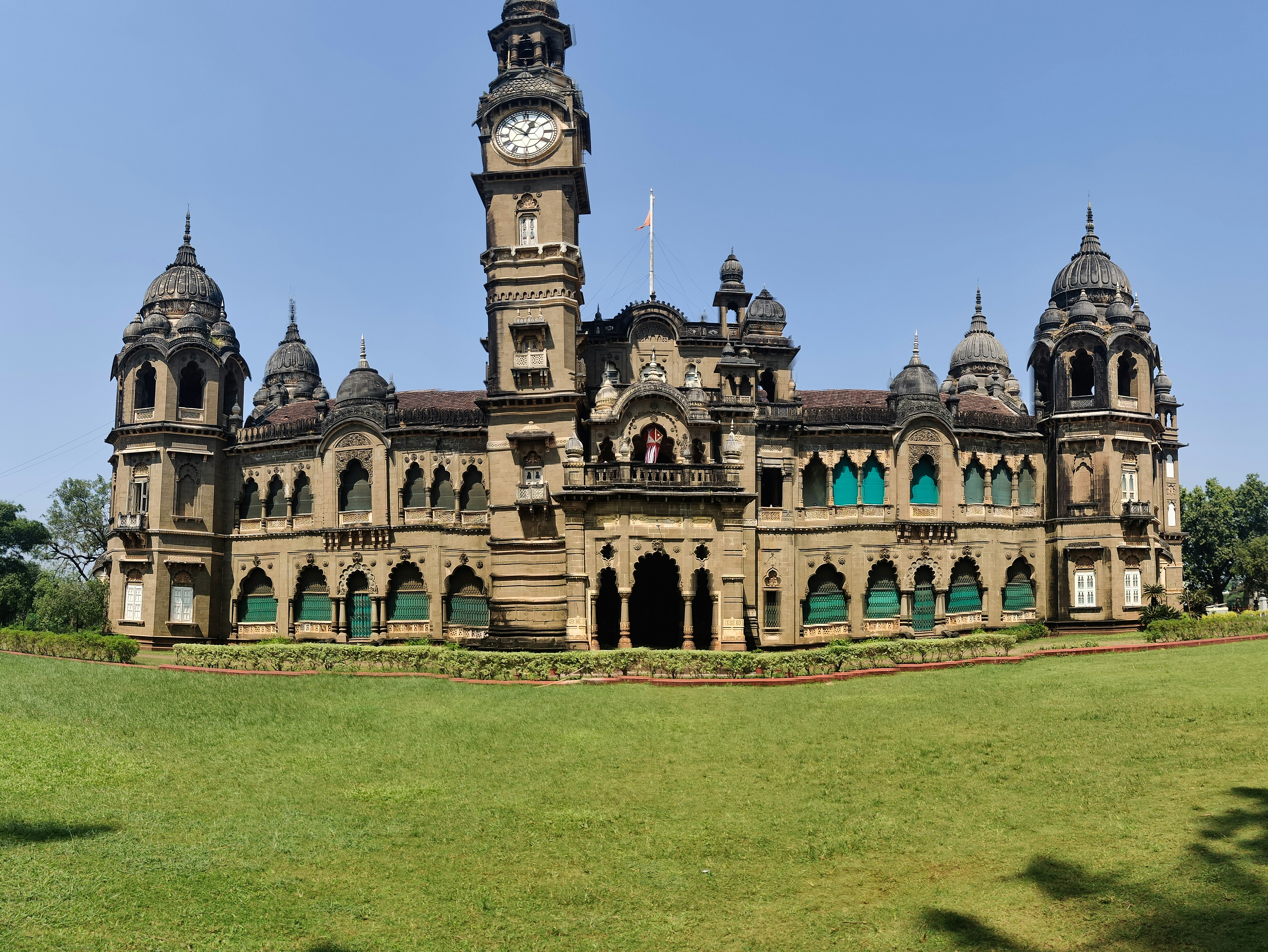 Ornate historic building with a central clock tower flanked by domed turrets, set against a clear blue sky with a manicured lawn in the foreground.