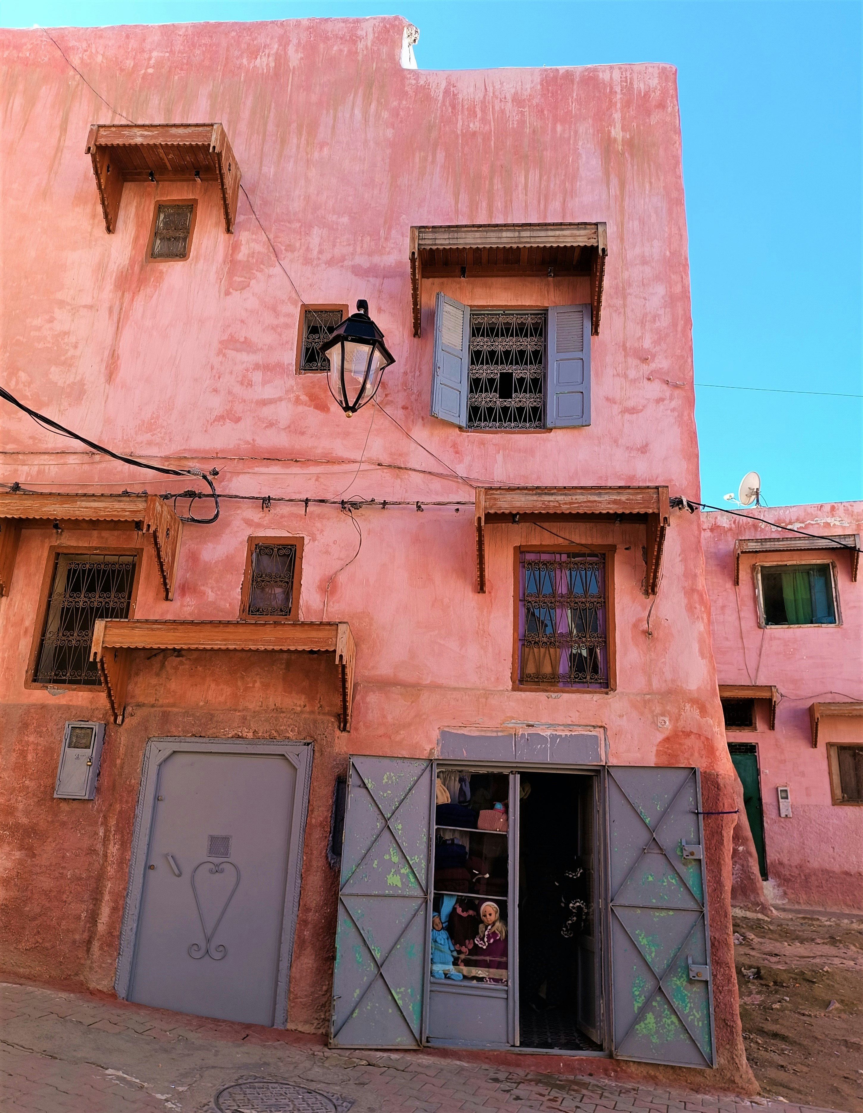 Photograph of a sunlit pink adobe building with a hanging lantern, balconies, and open doors revealing a glimpse of people inside.