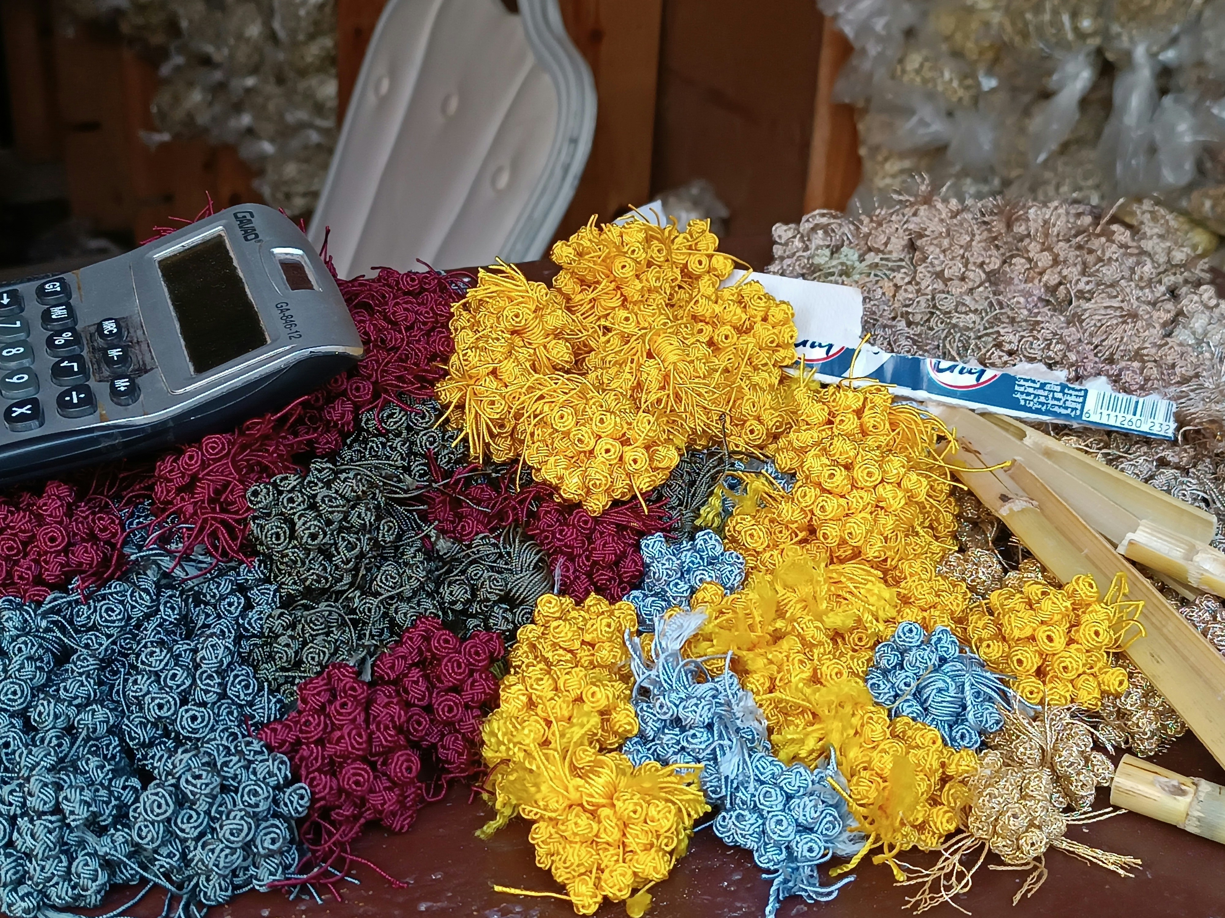 Colorful clusters of tiny paper flowers in blue, yellow, maroon, and beige spread across a workshop table, with a cordless phone and wooden sticks nearby.