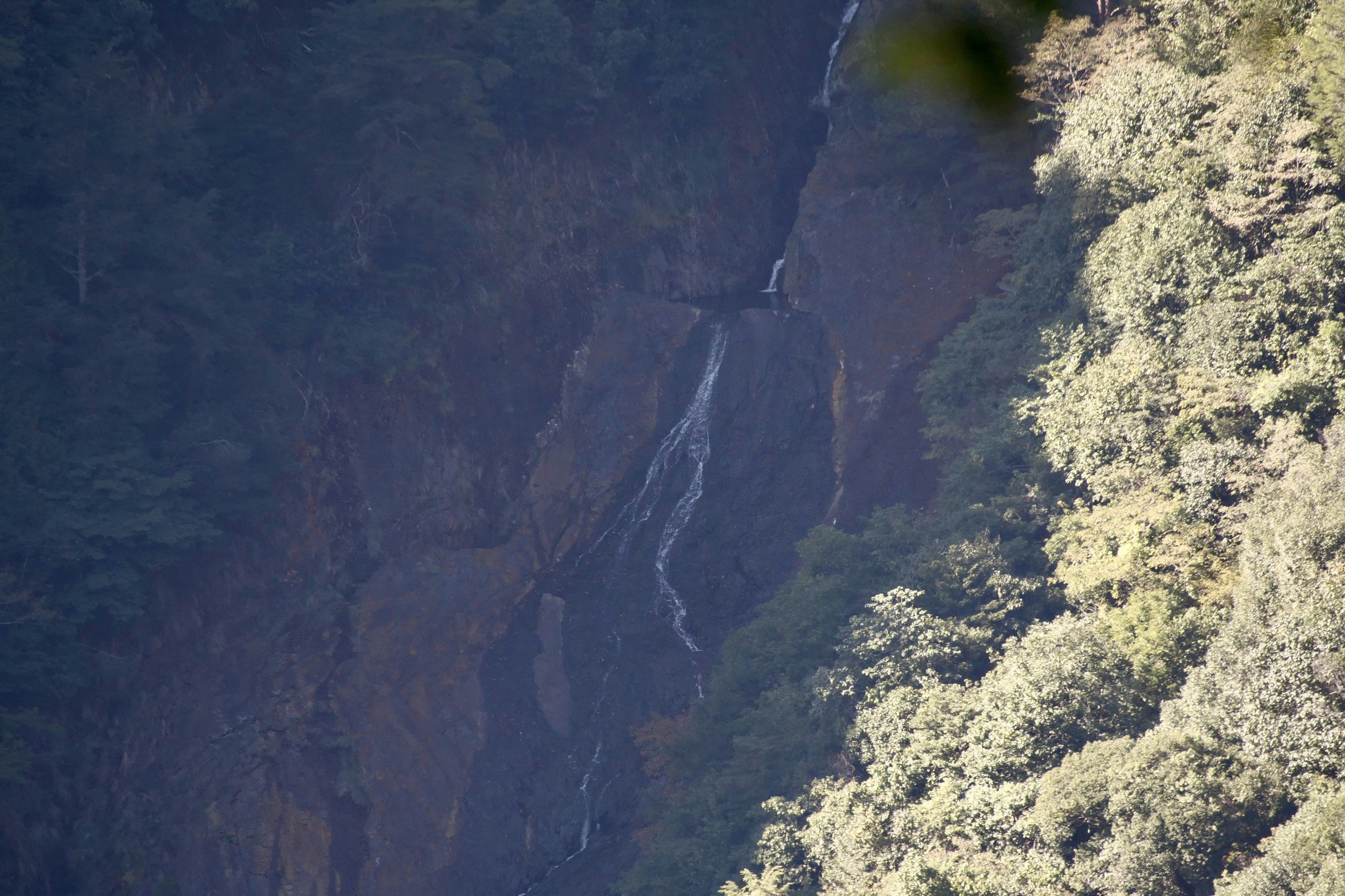 A slender waterfall threads down a rugged cliff face, with sunlit trees lining the right edge. A cool blue haze fills the left side, enhancing depth.
