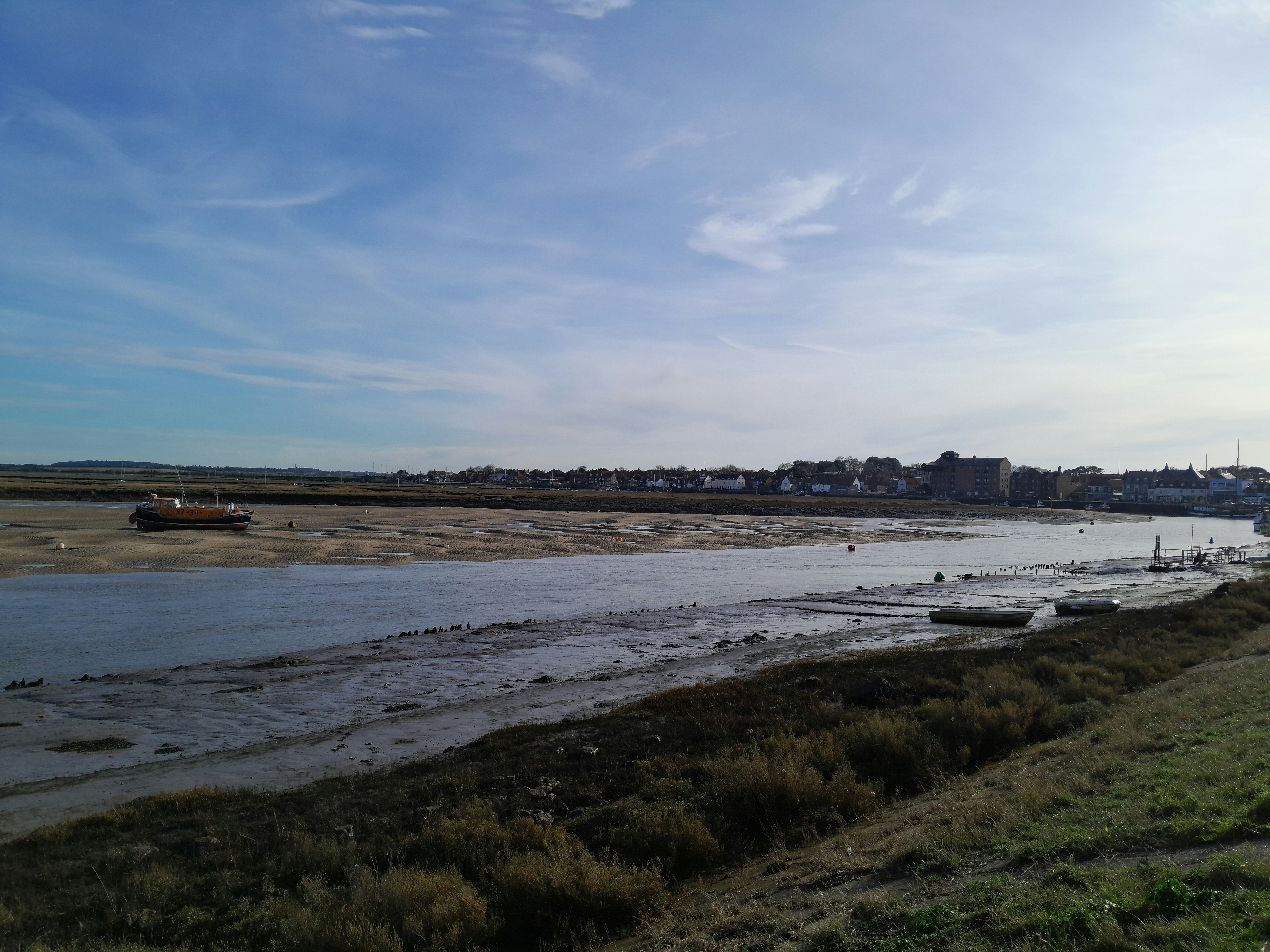 Calm coastal photograph of receding mudflats with a lone boat on the left and a distant town along the horizon. The scene highlights shoreline textures against an expansive sky.