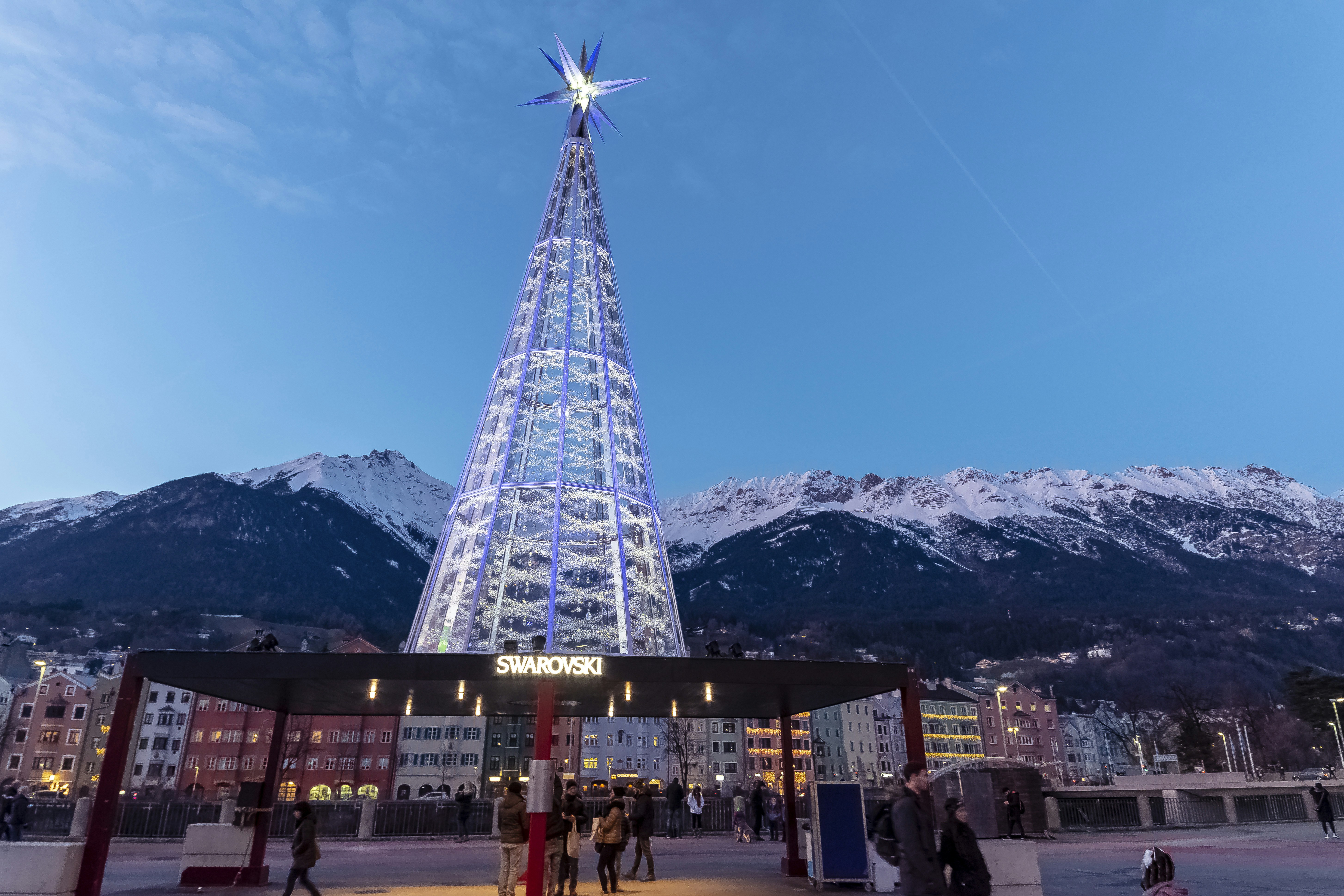 Night photograph of a luminescent crystal tower in a snowy alpine plaza, with Swarovski signage and pedestrians against a mountain backdrop.