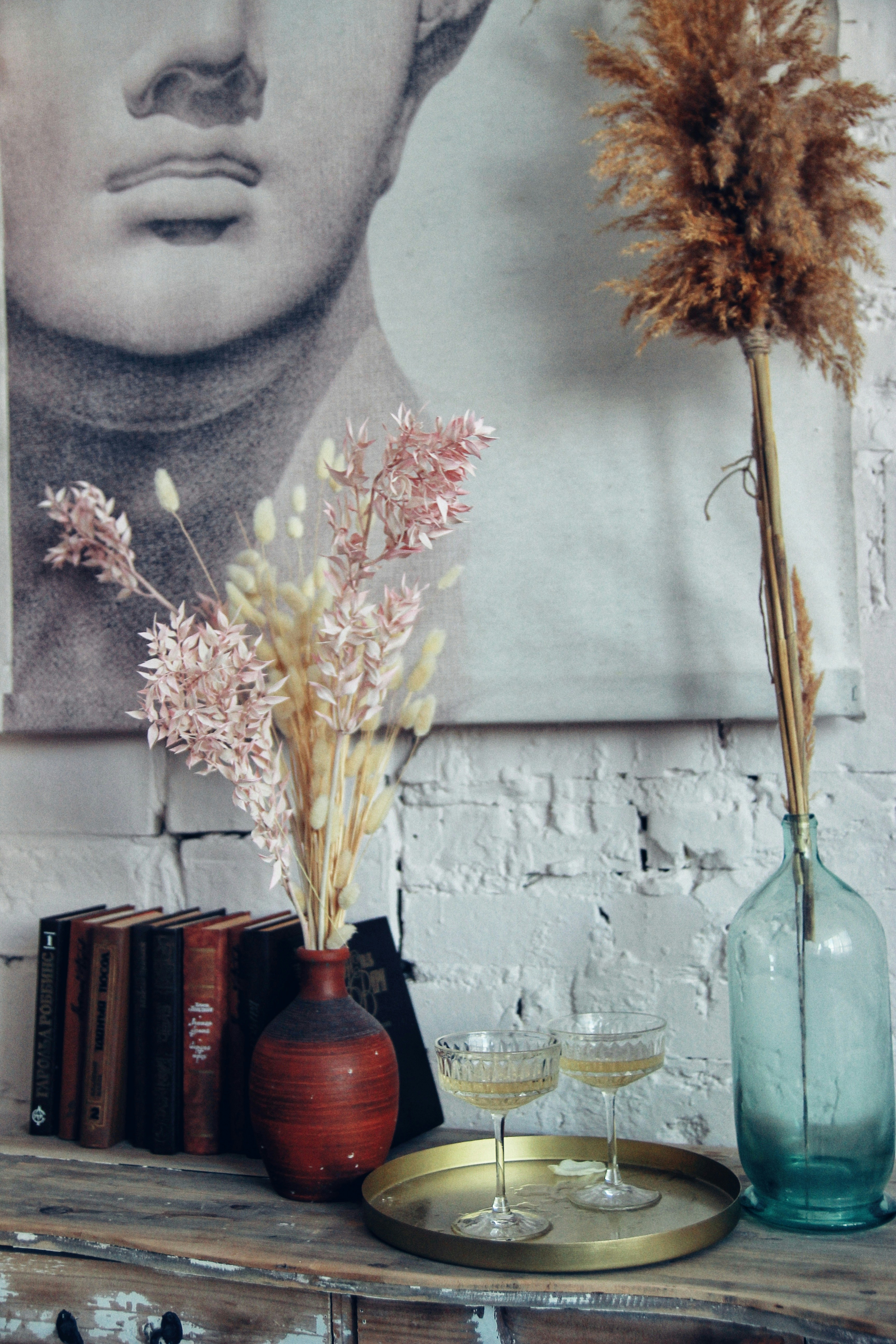 Delicate dried flowers in a vase beside two elegant glasses on a tray, with a monochrome portrait in the background. A blend of rustic and modern aesthetics.