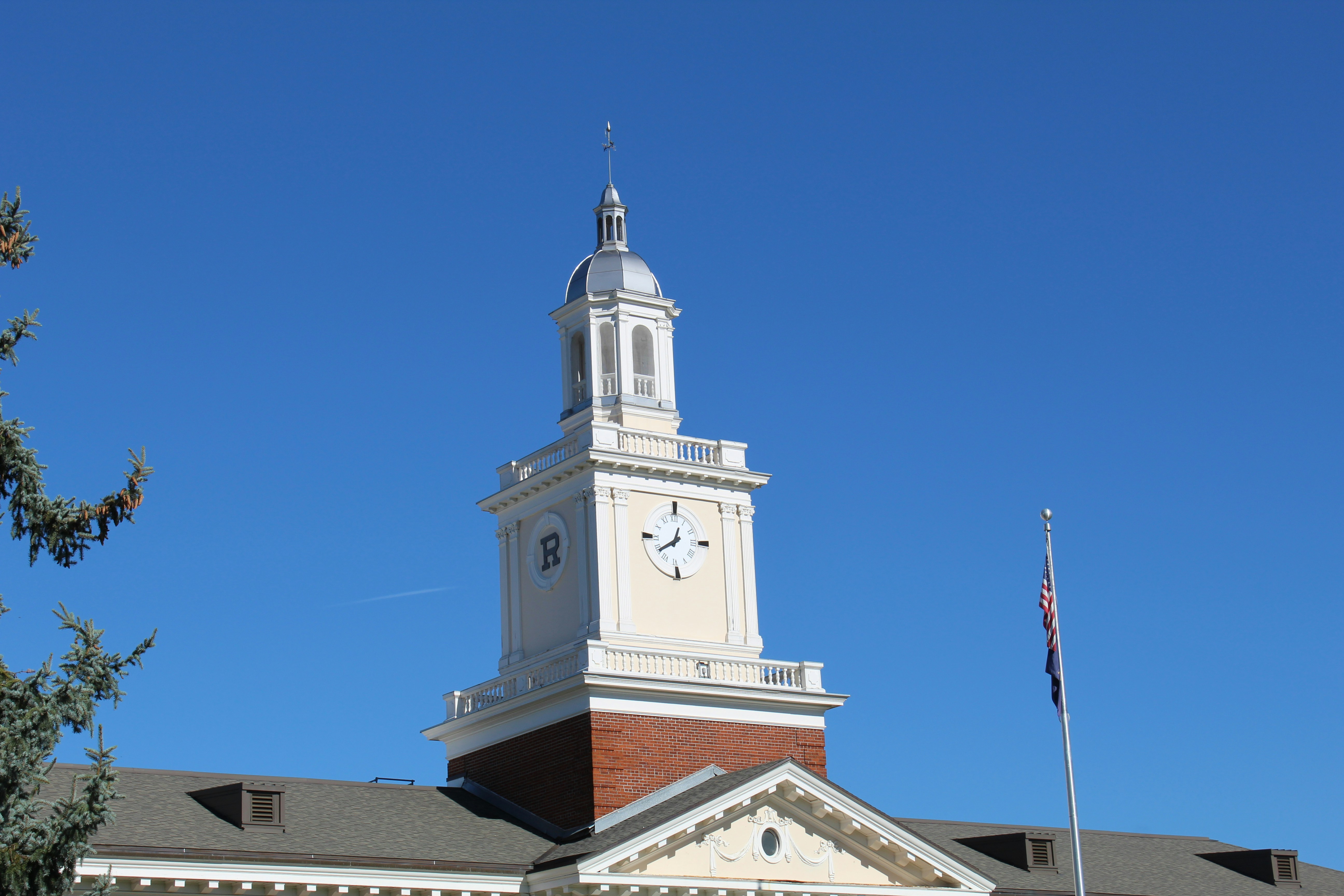 Clock tower of a school building under a clear blue sky with a tree and flagpole.