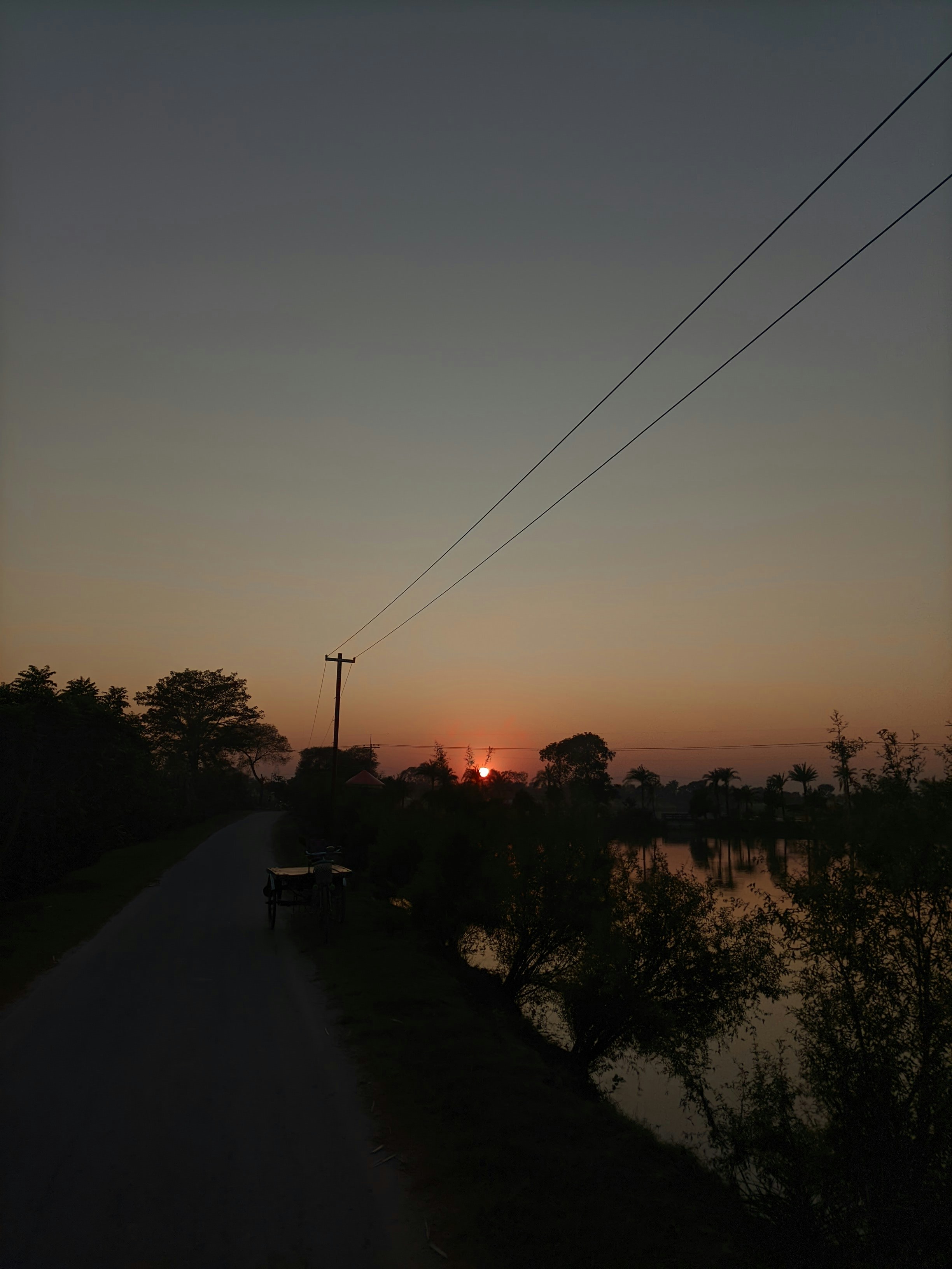 Sunset casting warm hues over a tranquil road beside a reflective water body, framed by silhouettes of trees and power lines.